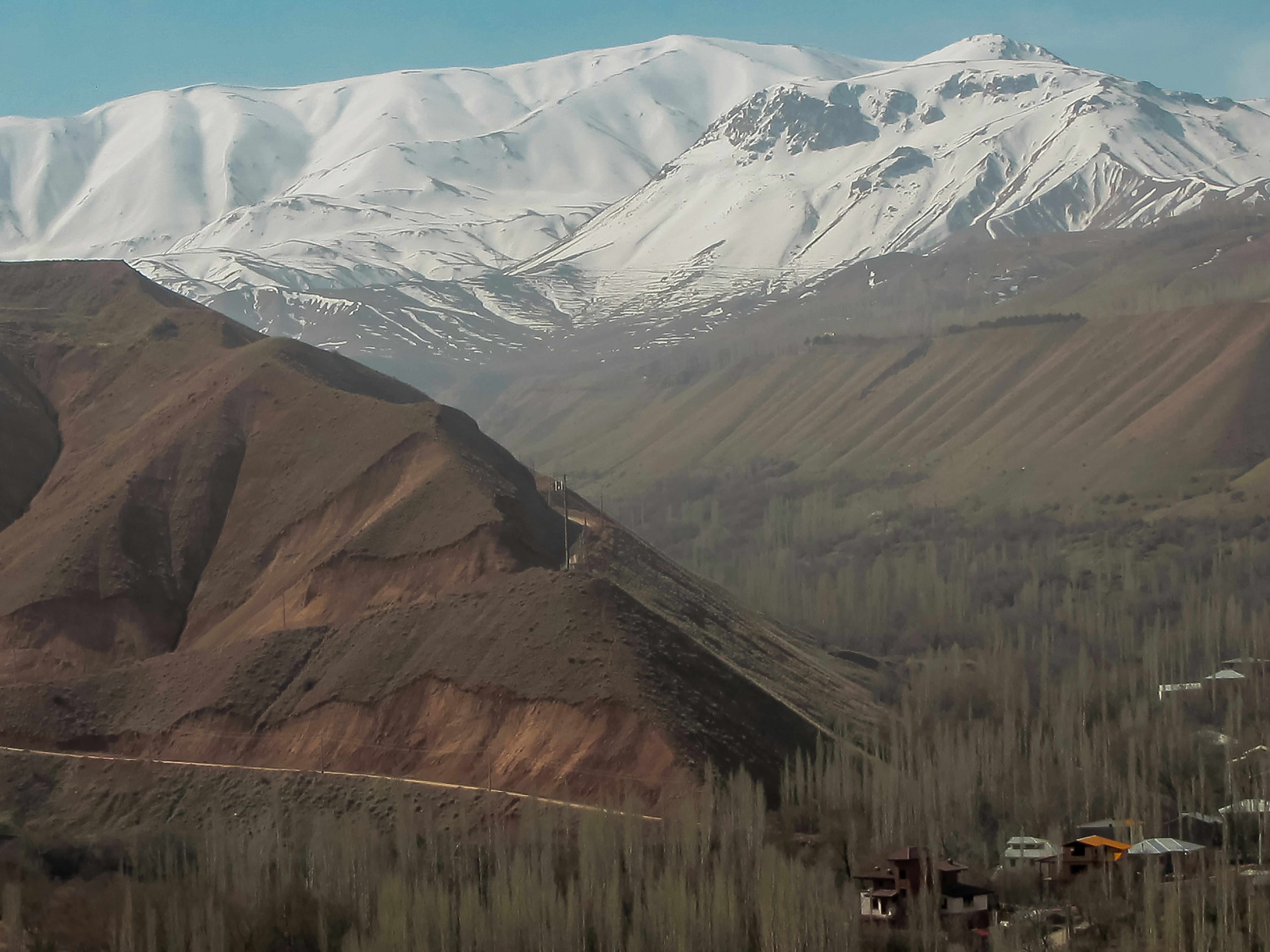 Snowy mountain backdrop rises behind rugged clay hills and a small settlement at their base, highlighting the contrast between natural grandeur and human presence.