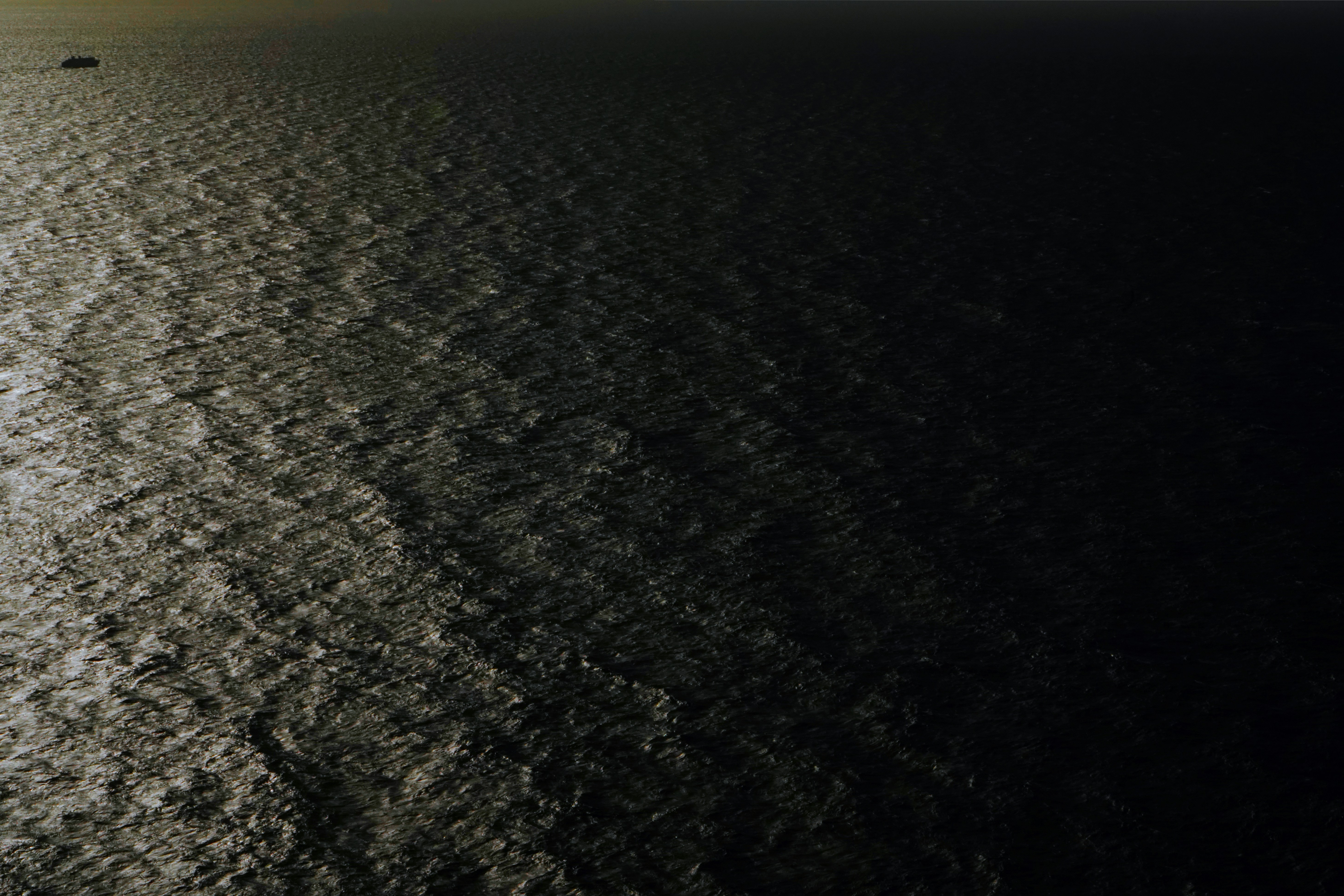 Glistening waves stretch across a tranquil sea, reflecting soft light and shadows. A solitary vessel drifts in the distance.