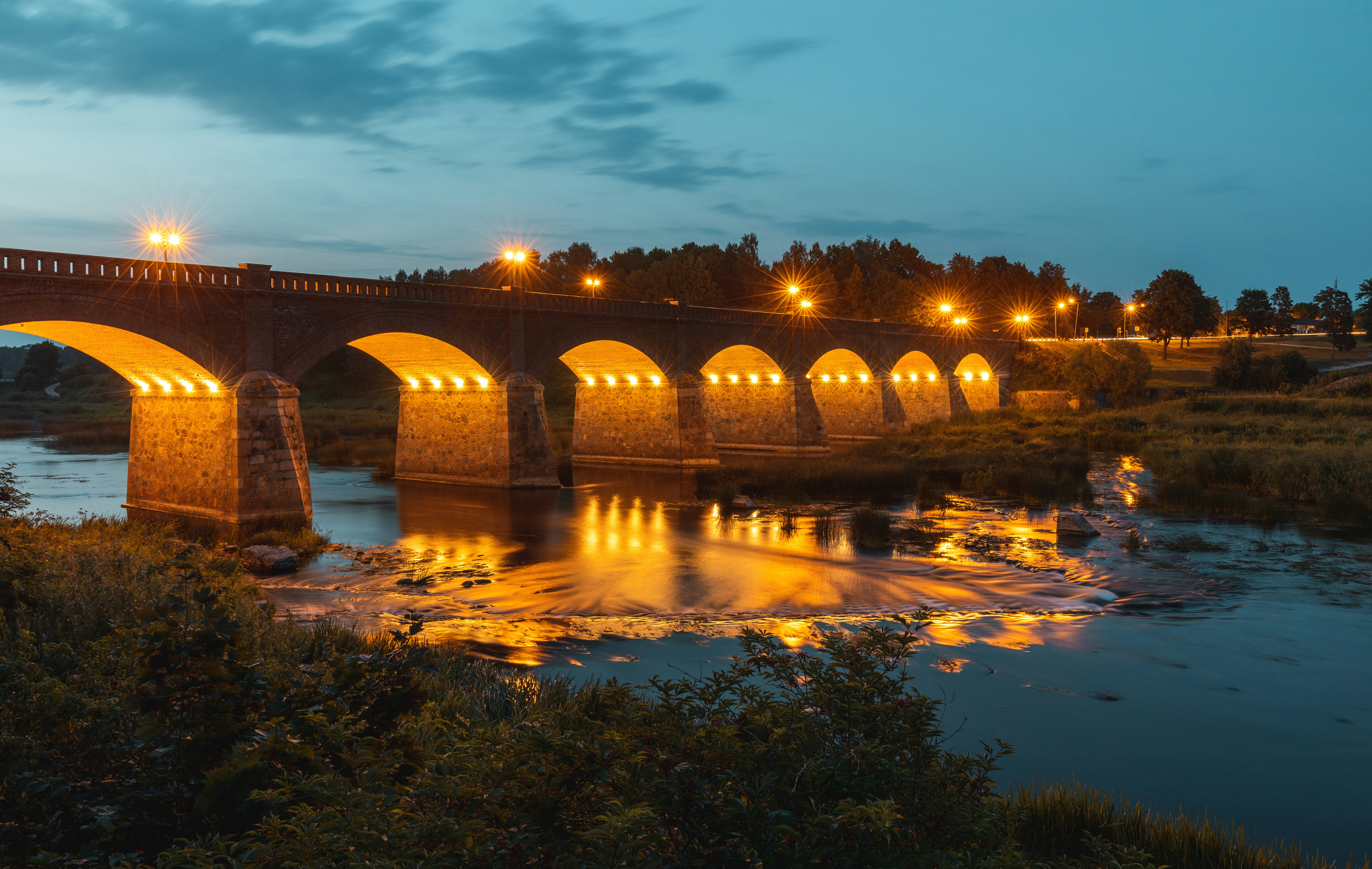 Illuminated bridge arches reflecting in a serene river under a twilight sky.