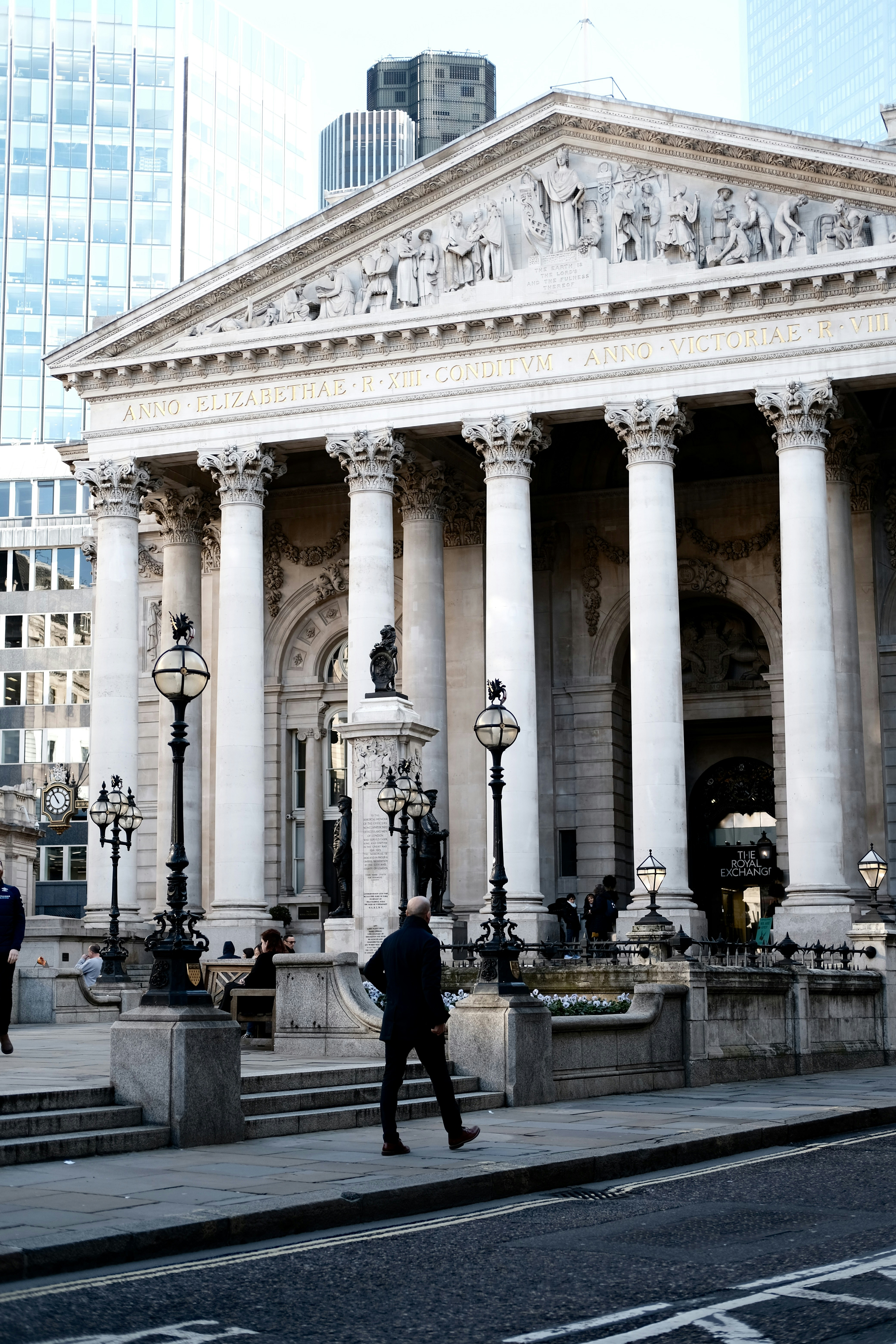 Classical building facade featuring grand columns and intricate sculptures, with pedestrians and street lamps adding life to the scene.