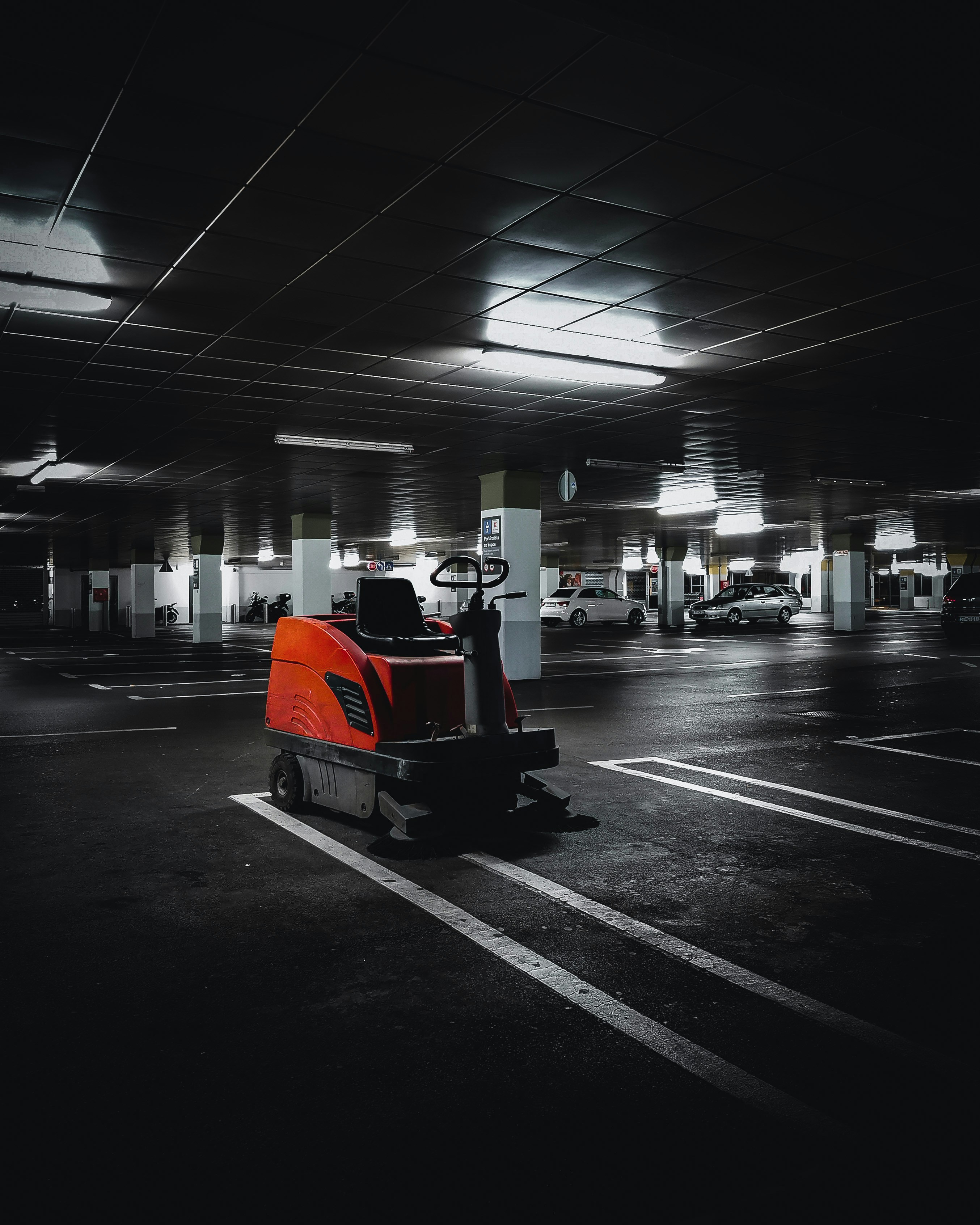A solitary floor cleaning machine stationed in a dimly lit parking garage, surrounded by empty parking spaces. The contrast between the vibrant red machine and the dark surroundings creates a striking visual.