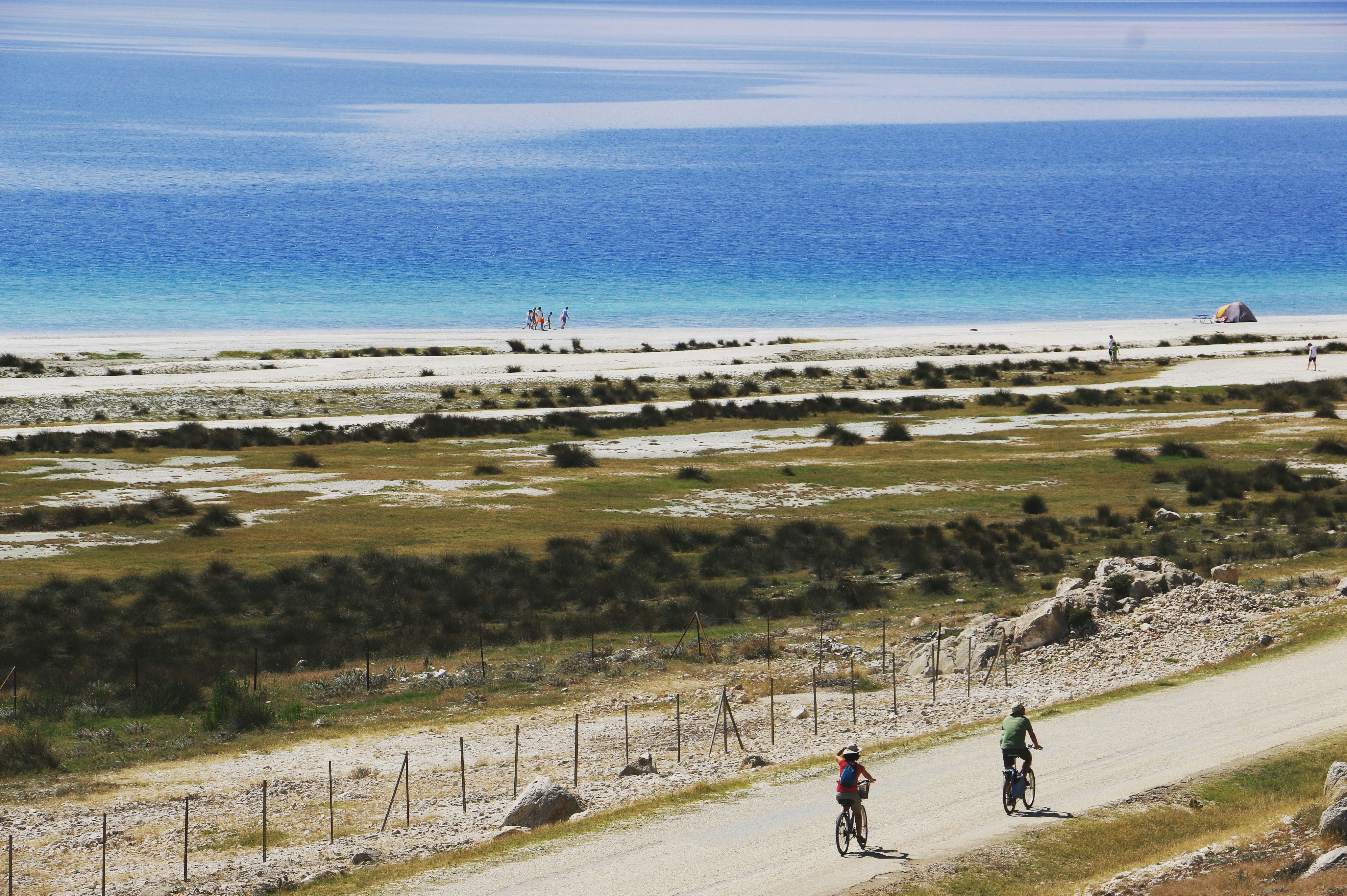 Two cyclists ride along a coastal path with vibrant blue sea and expansive sky in the background.