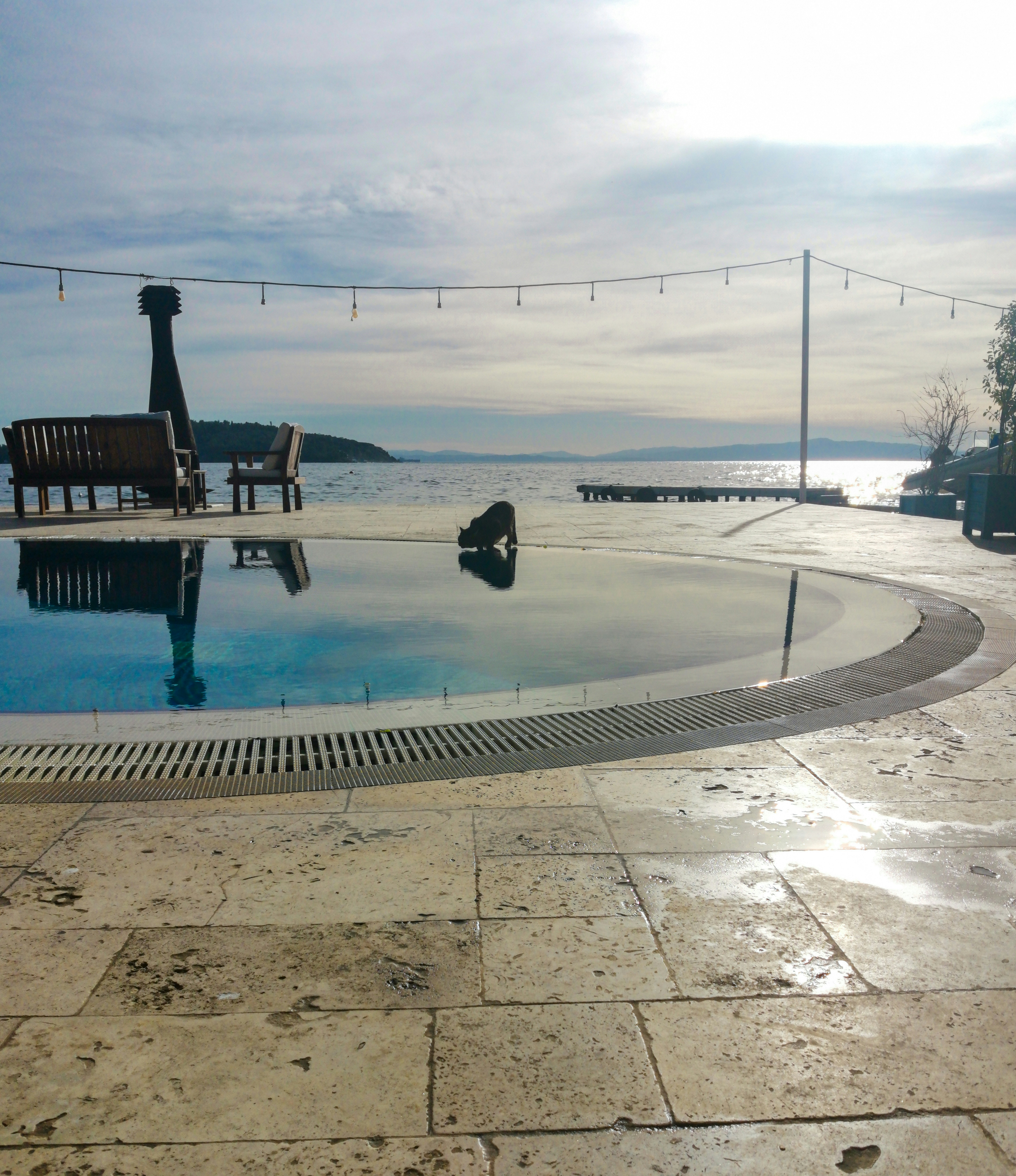 Tranquil poolside scene featuring a circular pool reflecting the sky, surrounded by lounge chairs and a distant view of the sea.