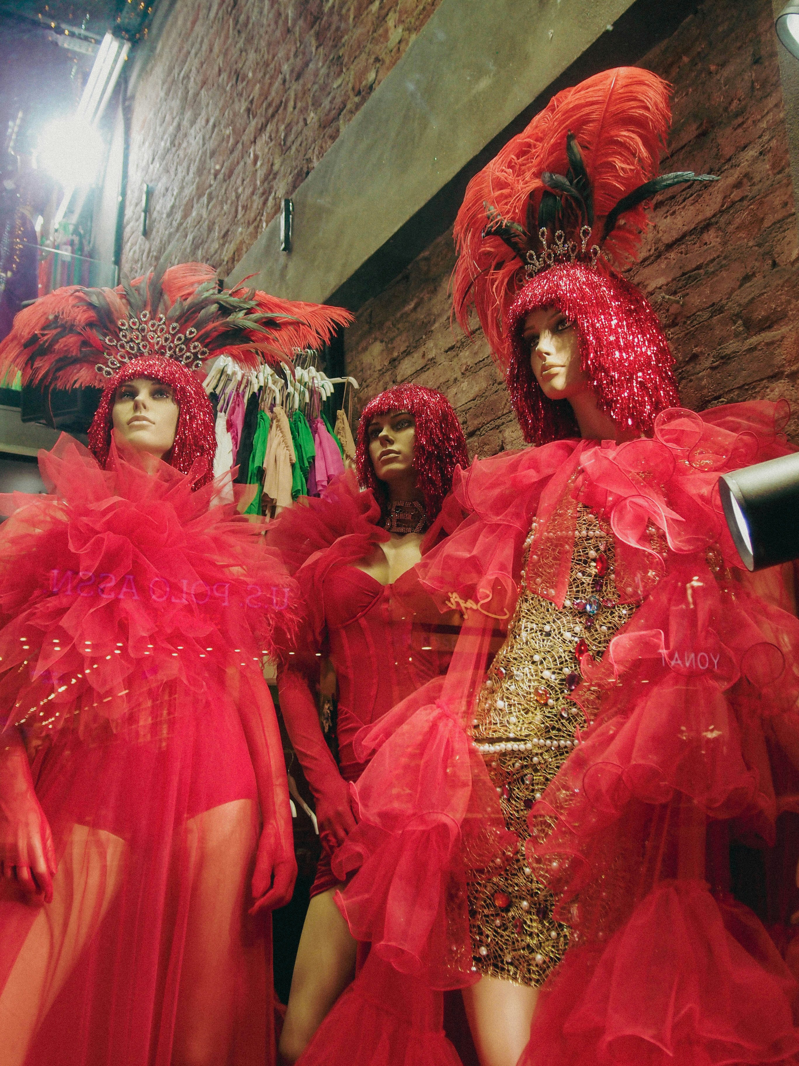 Mannequins in red feathered headdresses and sheer red gowns center a gold sequined dress in a shop window display. Dramatic couture and stage-like lighting highlight texture and fabric movement.