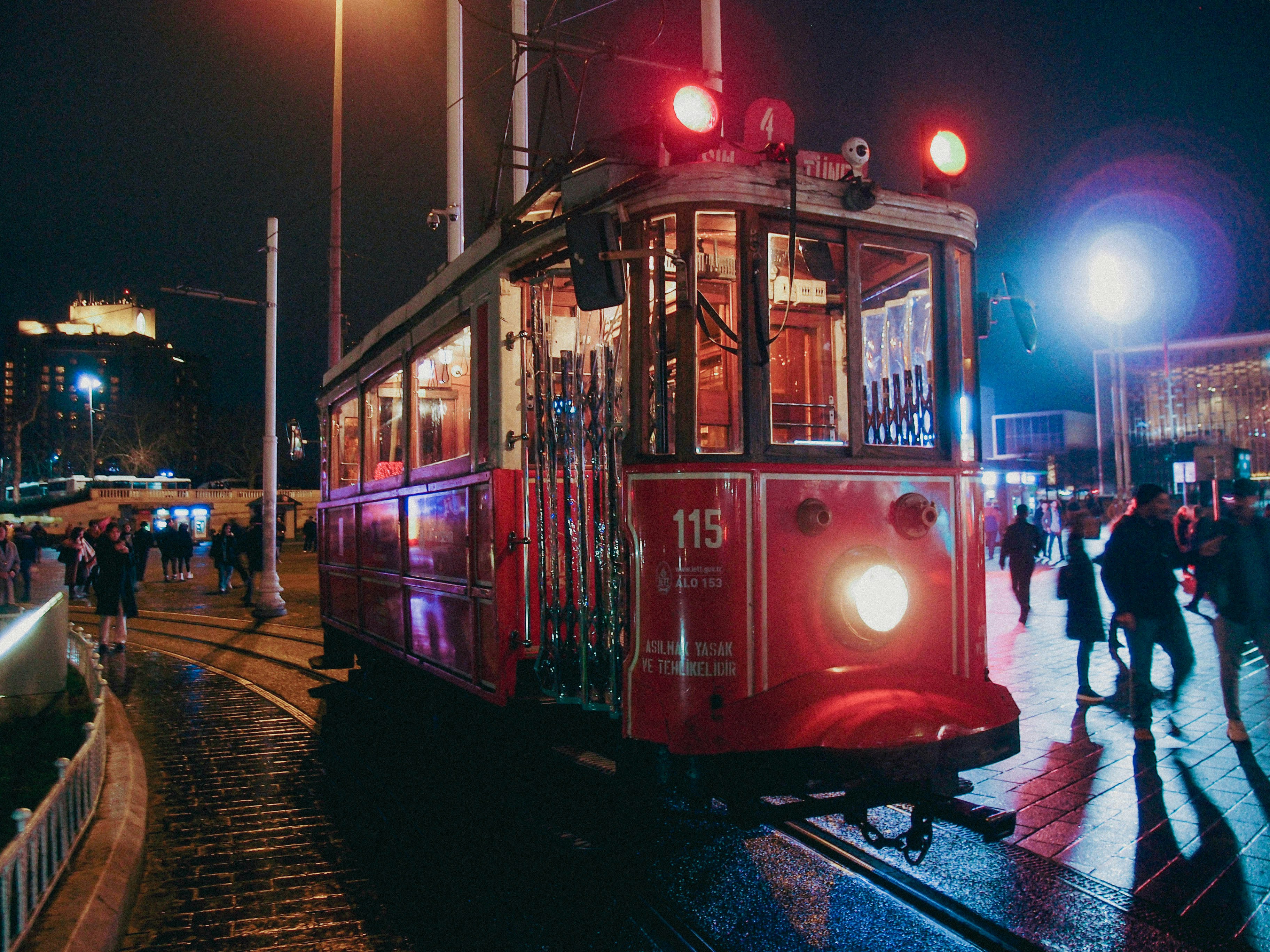 A vintage red tram illuminated by bright lights, surrounded by pedestrians in a bustling urban setting at night.