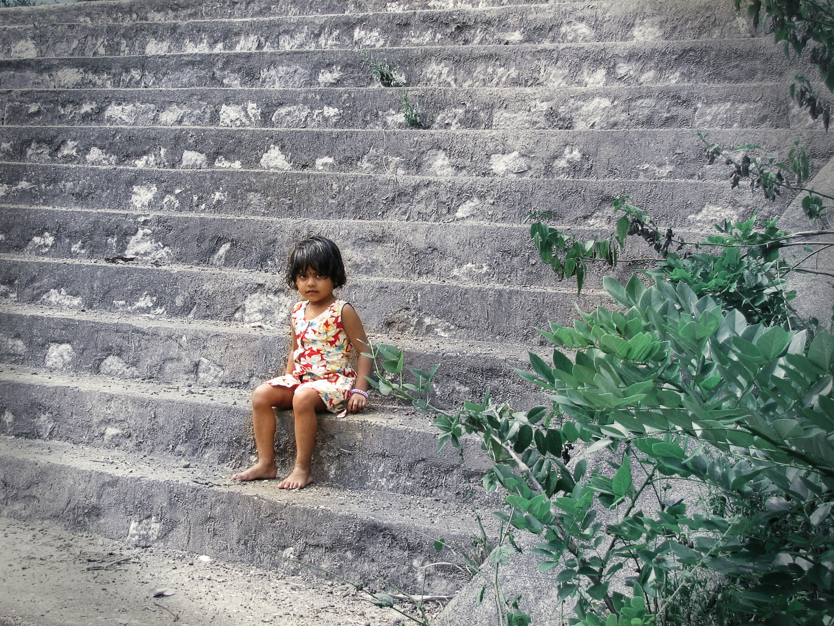 Young child sitting on weathered stone steps surrounded by greenery. The scene captures a quiet, contemplative moment in nature.