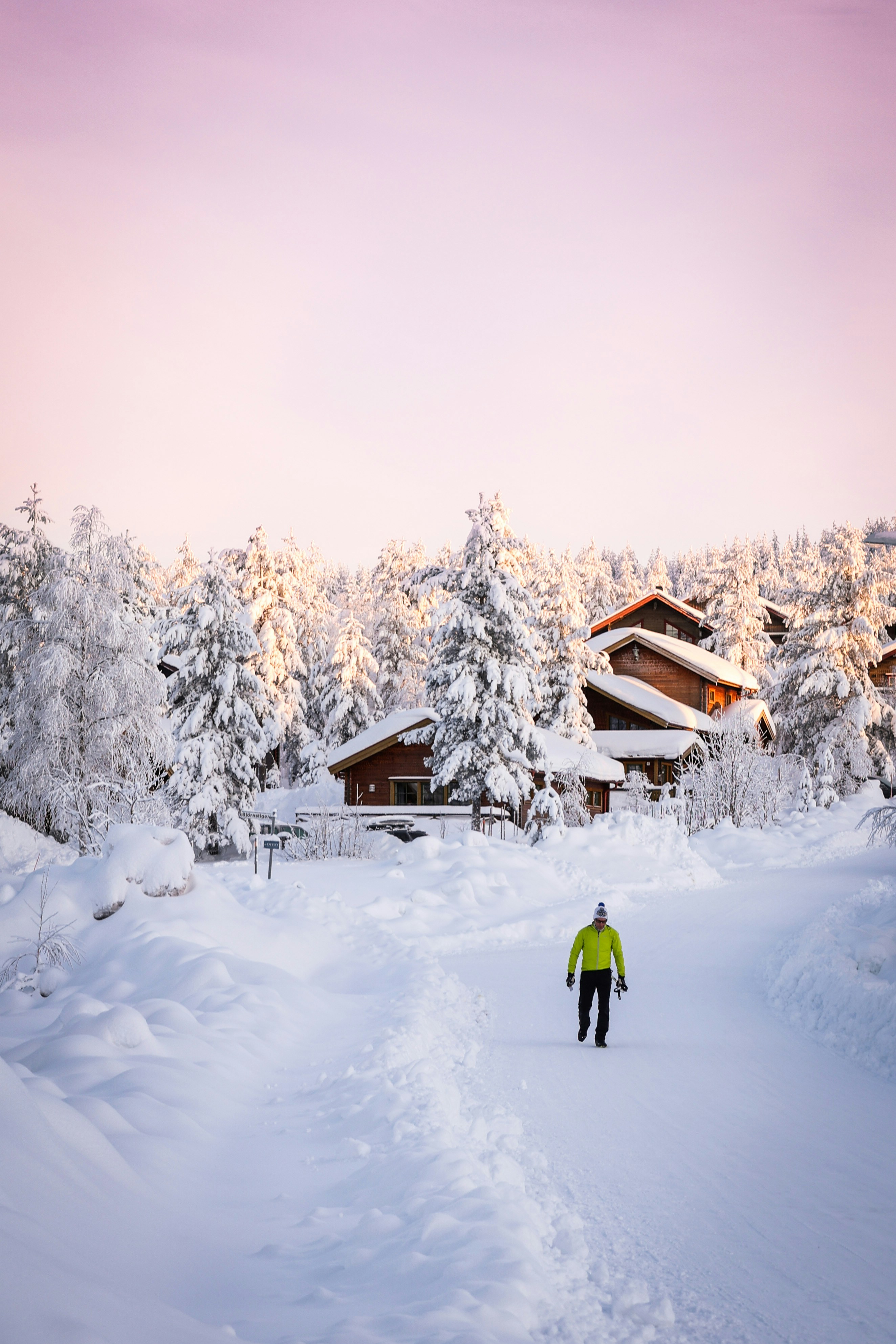 A lone figure walks along a snow-blanketed path, flanked by frosted trees and a cozy cabin under a pastel sky.
