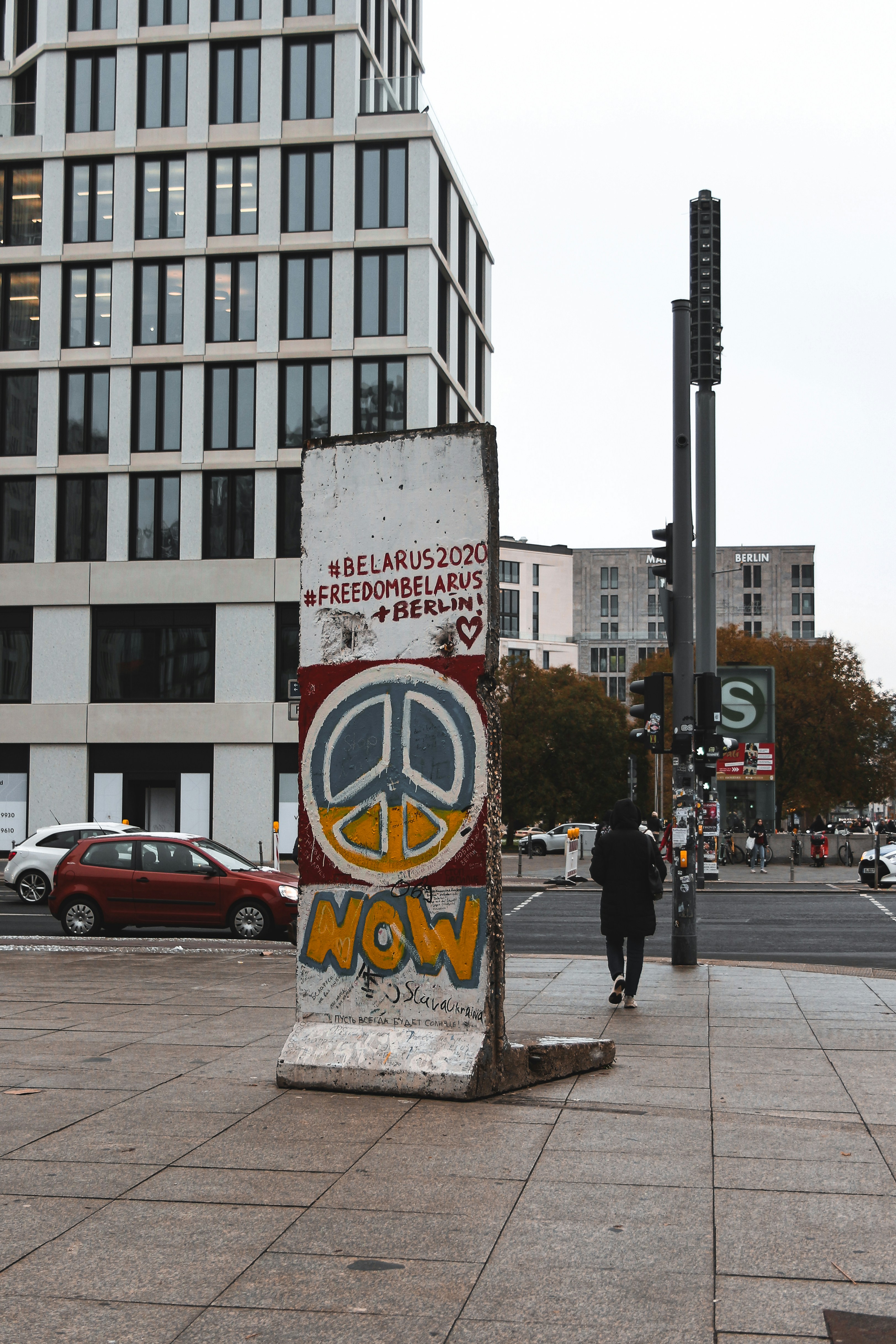 A segment of the Berlin Wall adorned with a peace symbol and hashtags advocating for Belarusian freedom. The urban backdrop contrasts the historical significance of the wall.