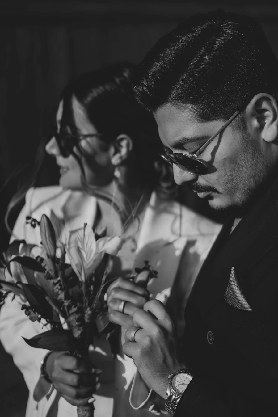 Black-and-white portrait of a man in sunglasses and a woman holding a bouquet, both lit by soft directional light. The man looks downward in the foreground while the woman stands slightly behind him.