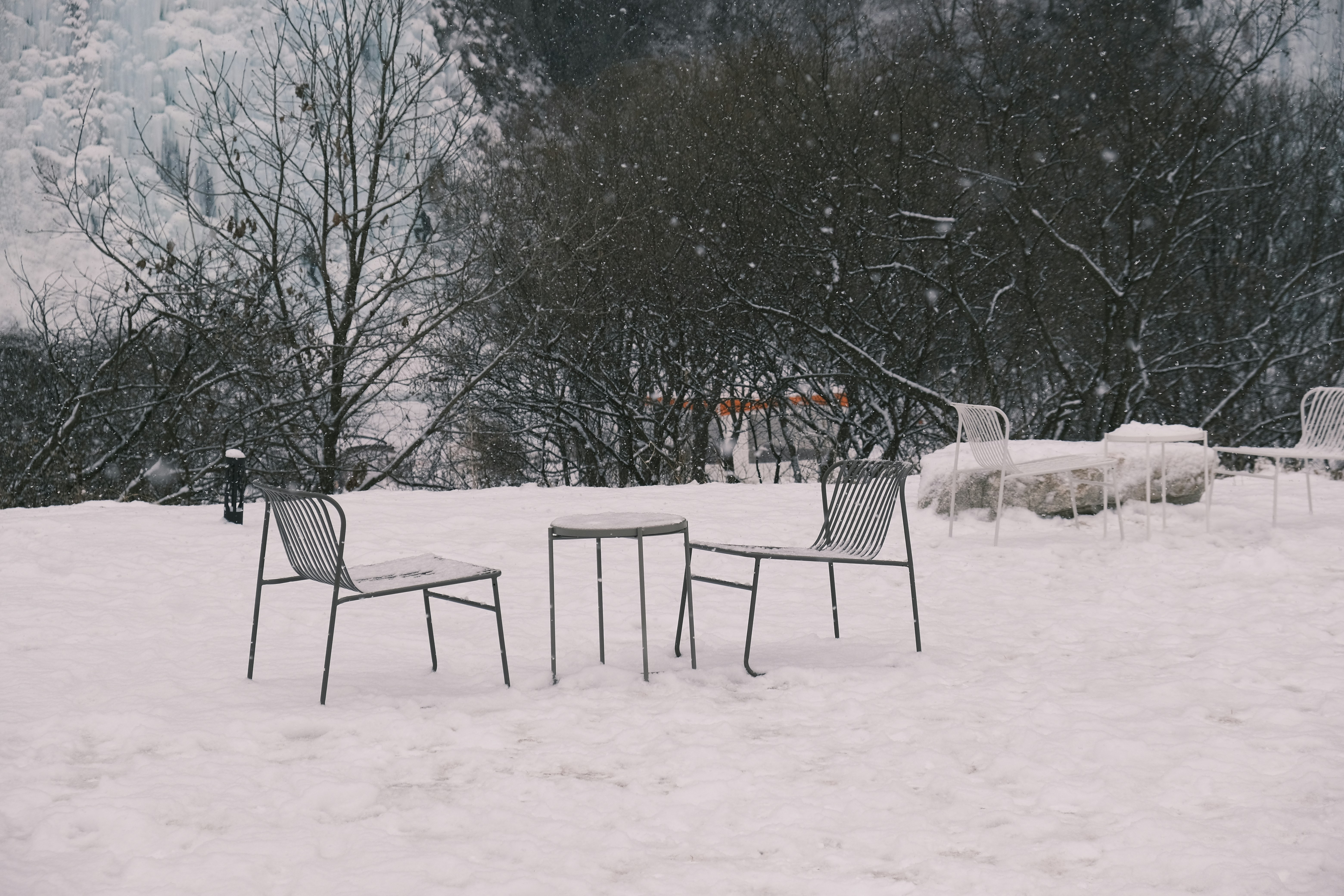 Two metal chairs and a table covered in snow, set against a backdrop of snow-laden trees.
