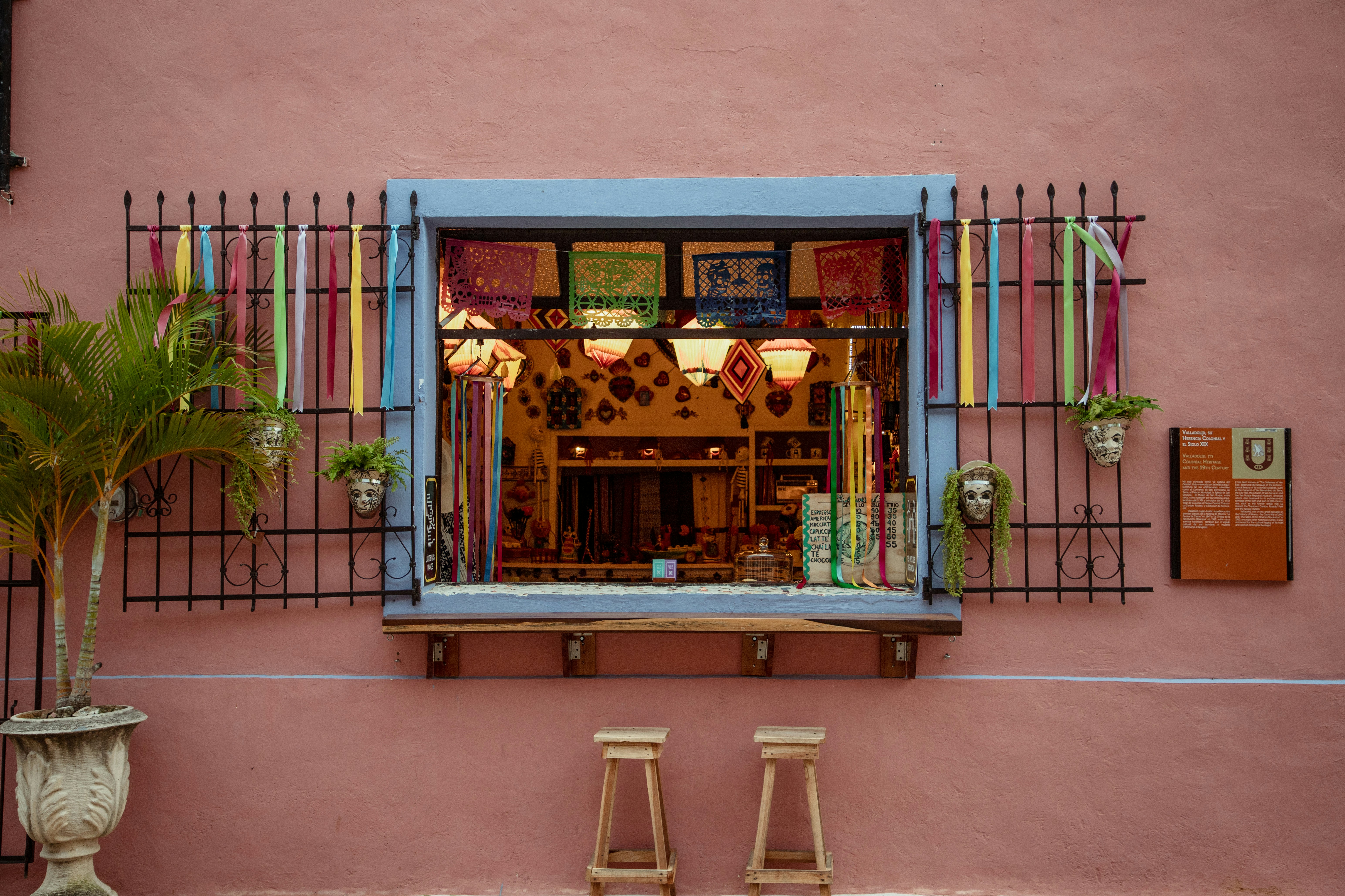 Colorful window display with plants and stools against a pink wall, evoking a cozy ambiance.