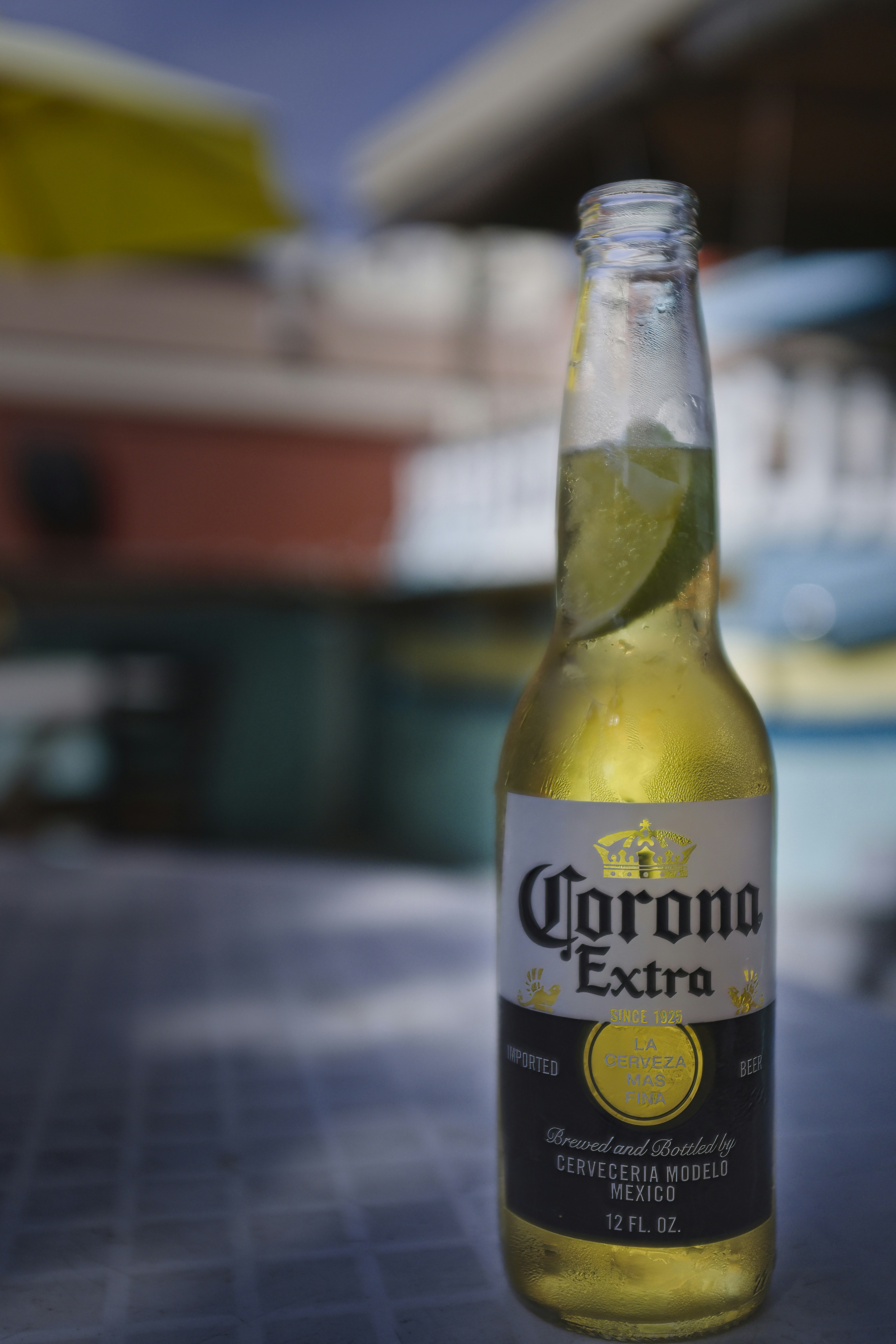 A Bottle of Corona Extra Beer on a Table Outside - A bottle of Corona Extra beer sits on a table outside. The bottle is partially filled with the golden liquid, and there is a lime wedge inside. The background is blurred and out of focus, but it appears to be a sunny, outdoor setting.