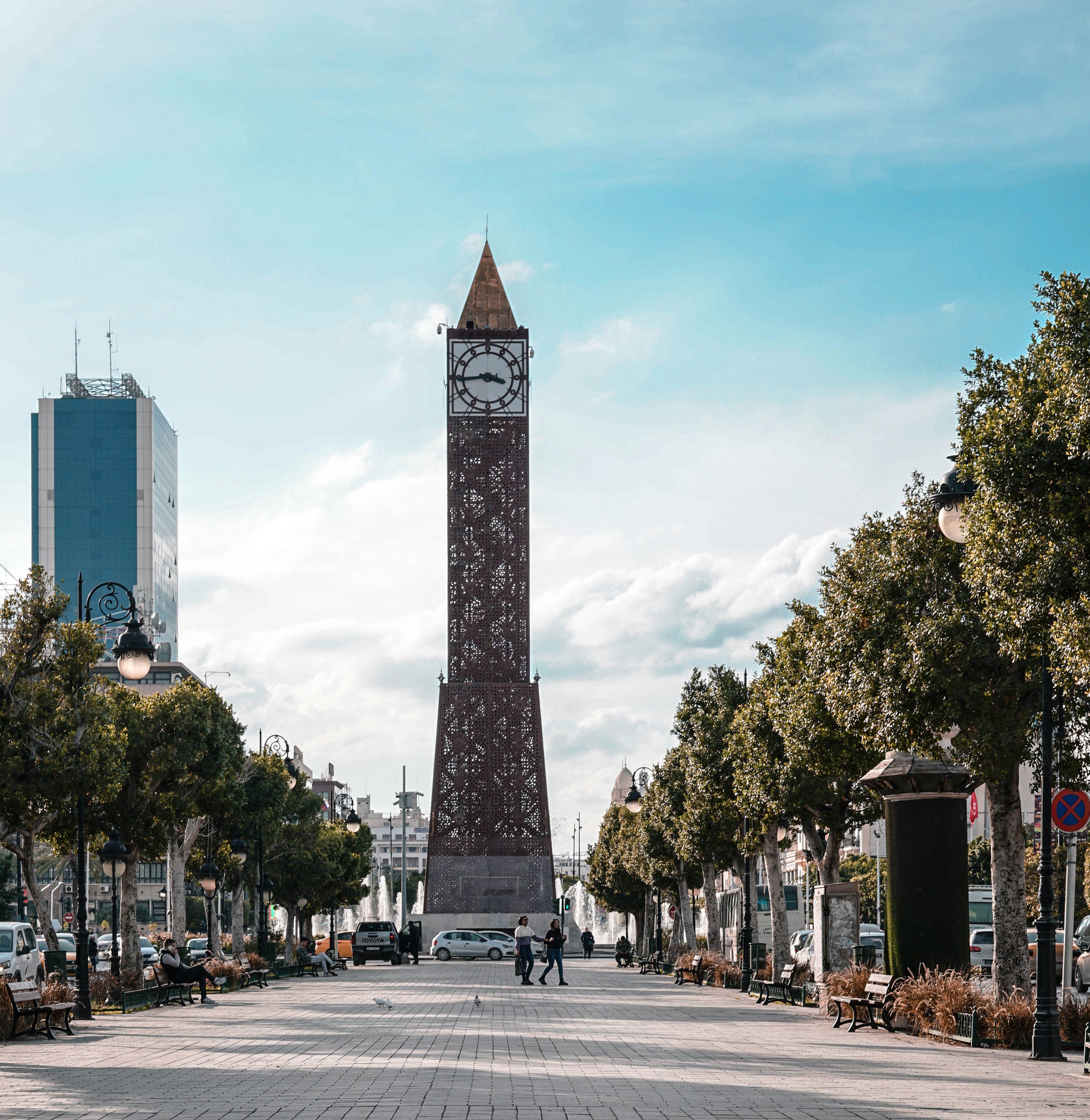 A striking clock tower stands prominently in a lively urban square, surrounded by trees and modern architecture. The scene captures the essence of city life and architectural beauty.