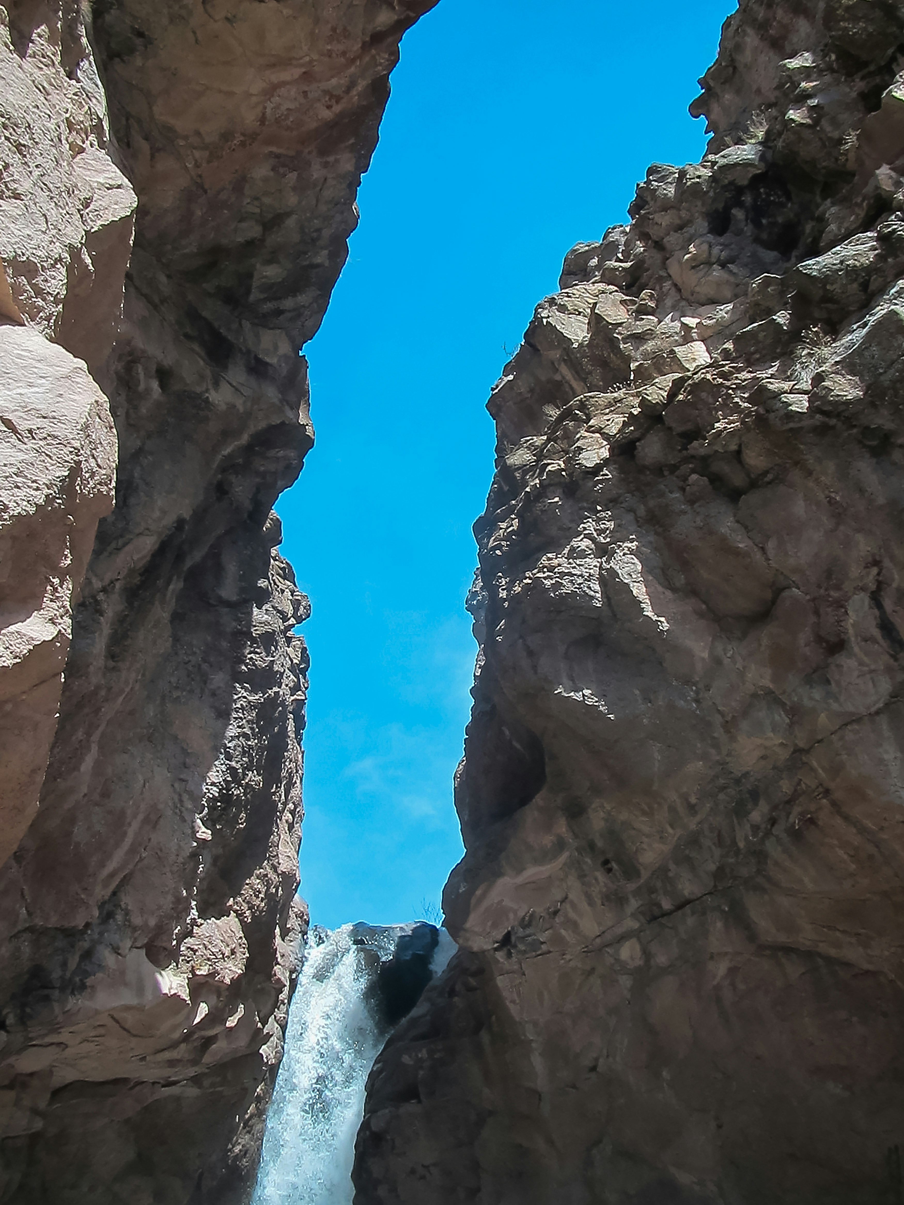 Tall canyon walls frame a bright blue sky with a thin waterfall at the base, emphasizing vertical scale in a narrow chasm.