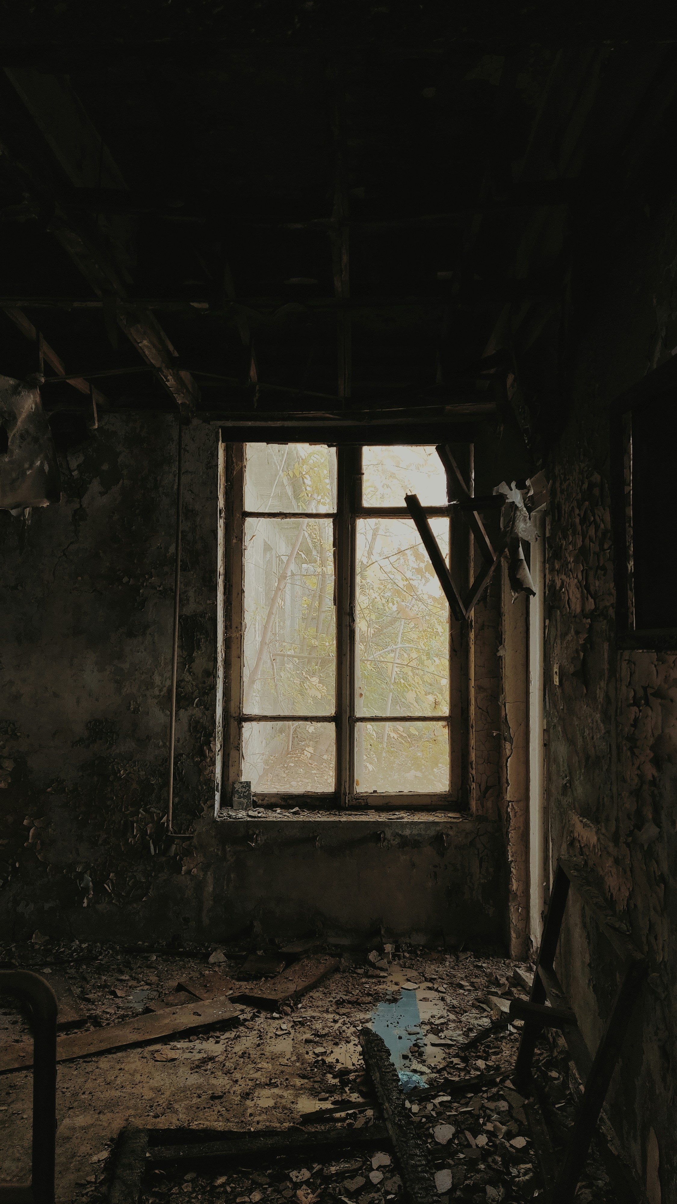 Decaying interior of a room featuring a weathered window framed by peeling walls and scattered debris. Sunlight filters through, hinting at the outside world.