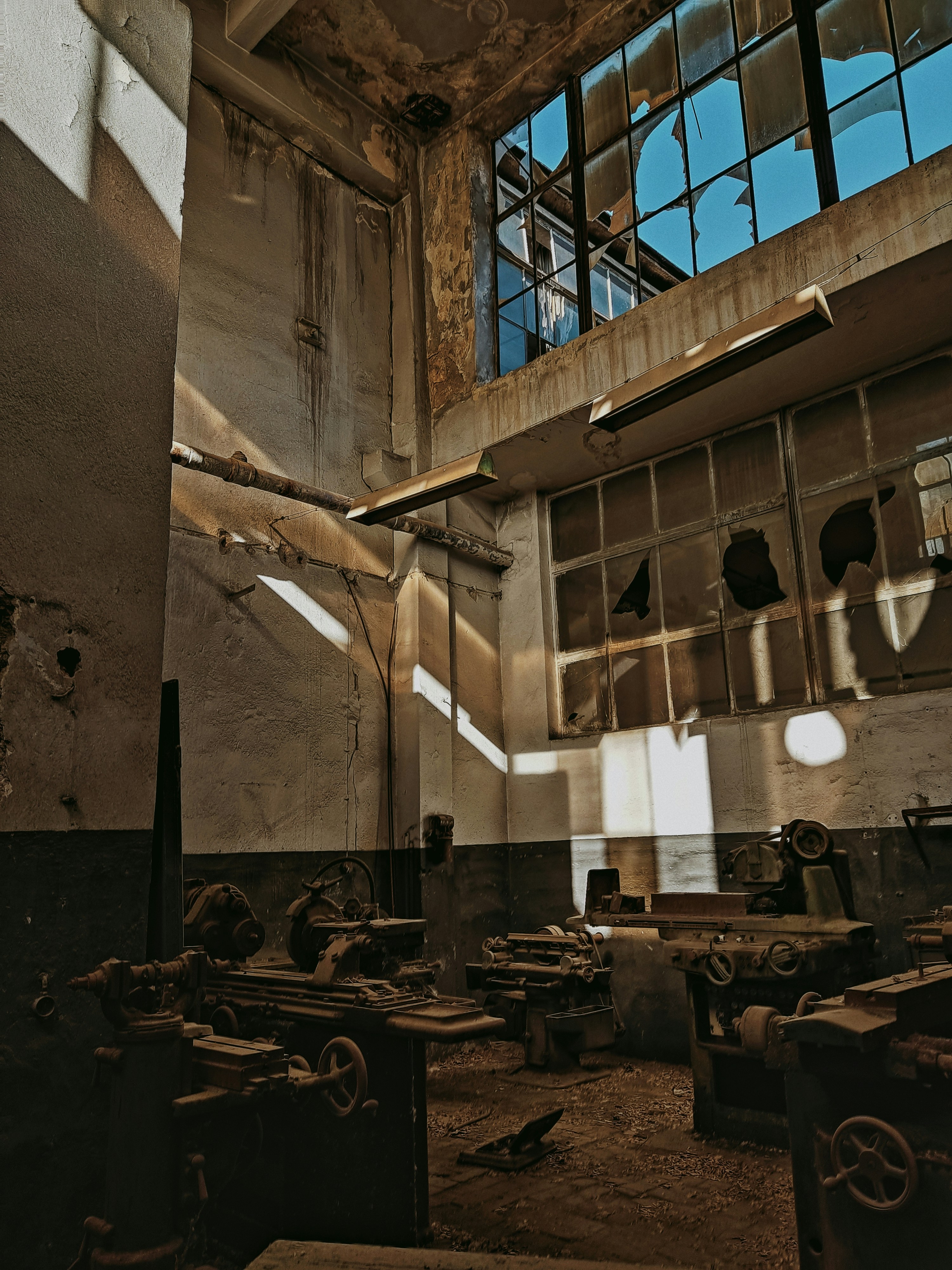 Sunlight filters through broken windows, casting shadows on rusted machinery in a derelict workshop. The atmosphere evokes a sense of forgotten industry.