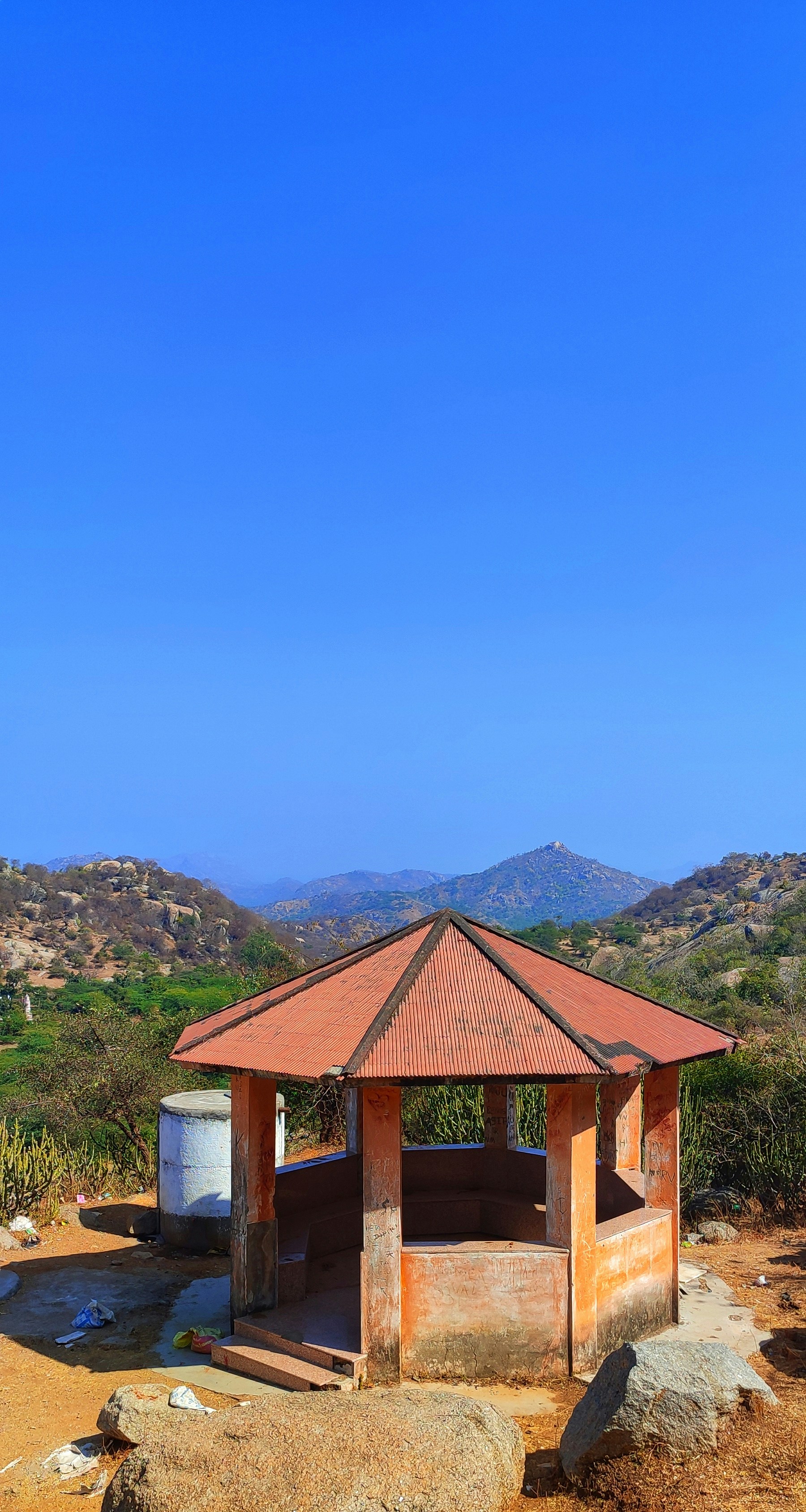 A rustic gazebo surrounded by lush greenery and rocky terrain under a clear blue sky.