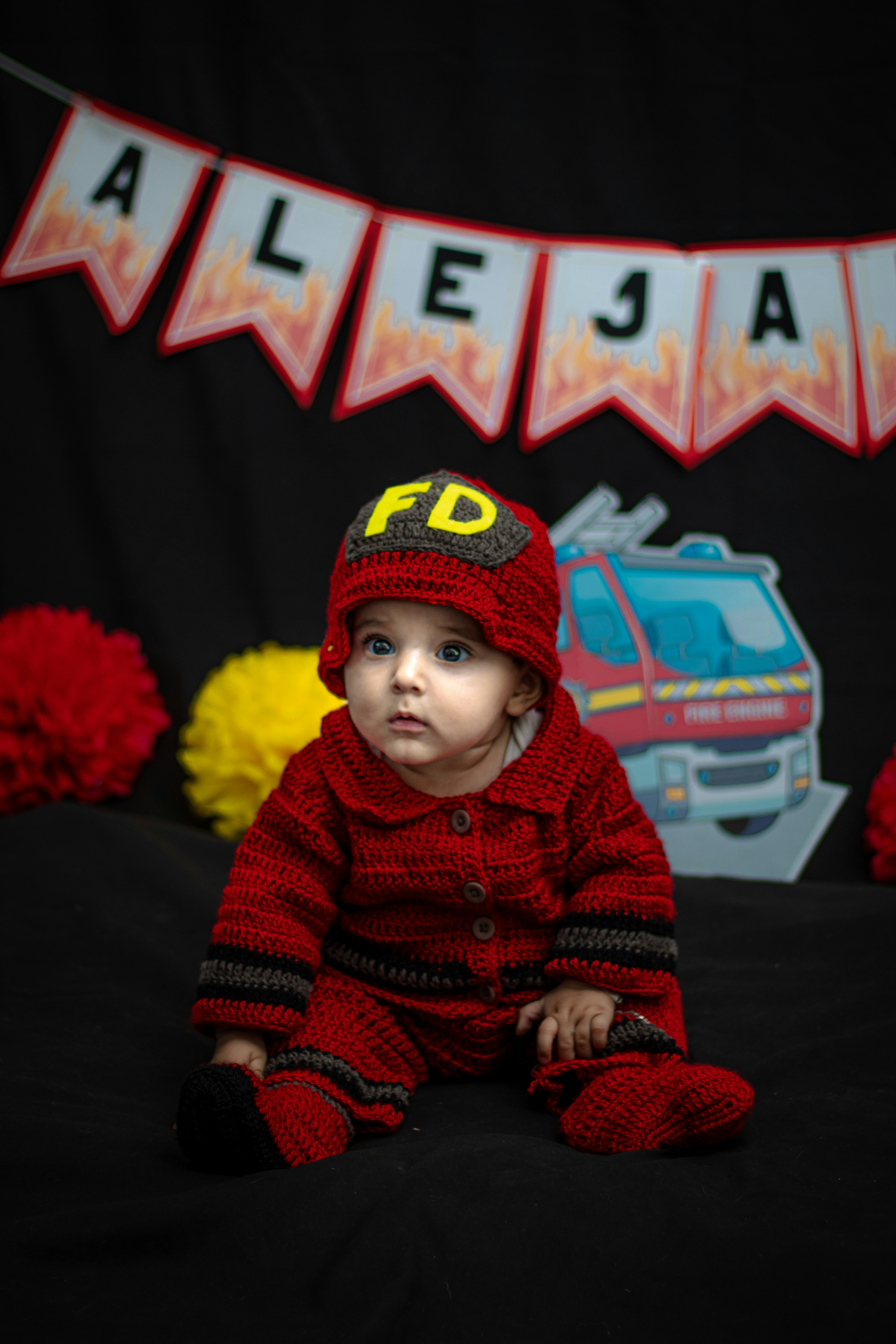 Tiny Firefighter in Knits: Portrait Beneath a Festive Banner and Cartoon Fire Truck