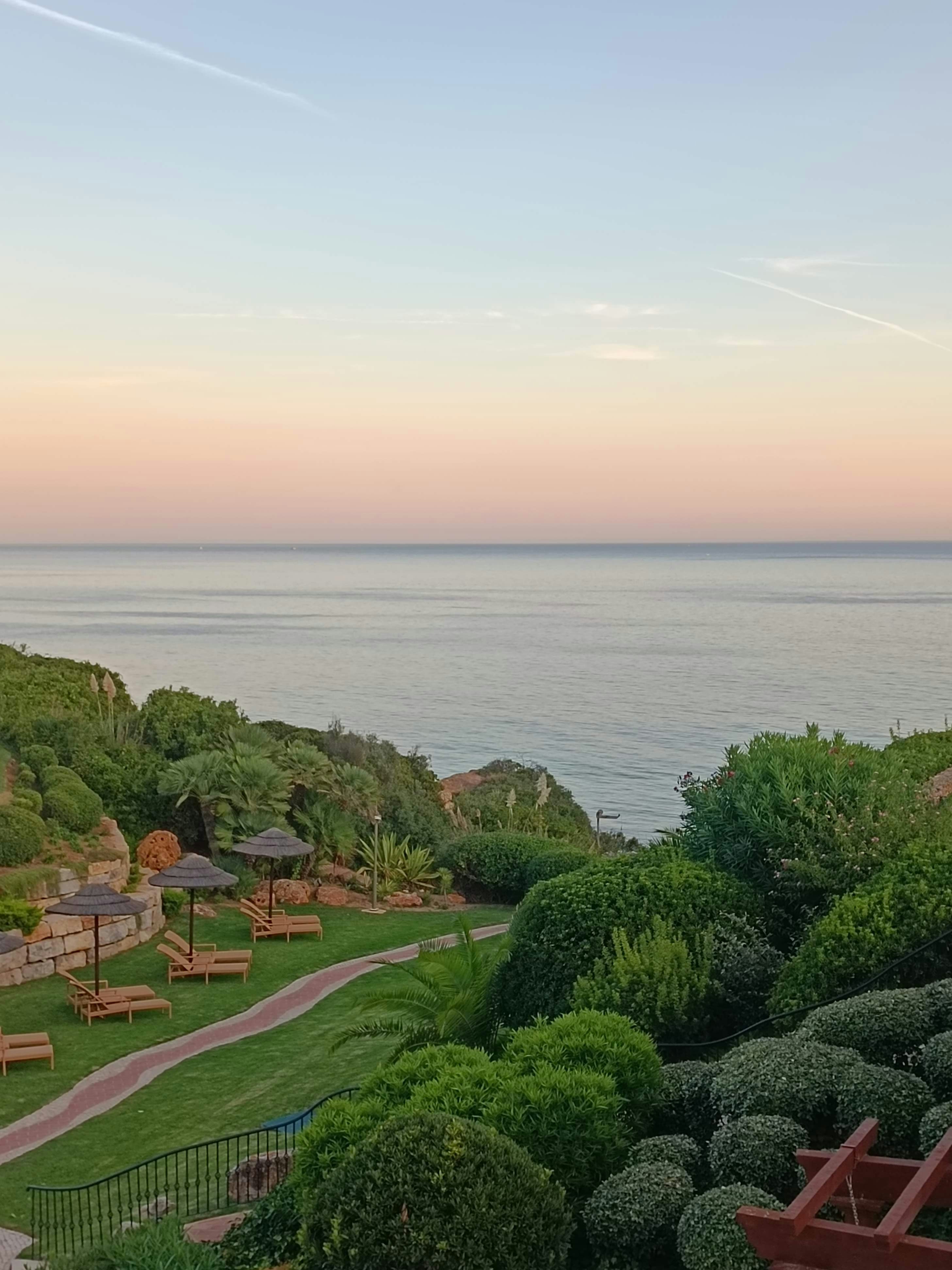 Lush garden with manicured hedges and wooden lounge chairs overlooking a tranquil sea at dusk.