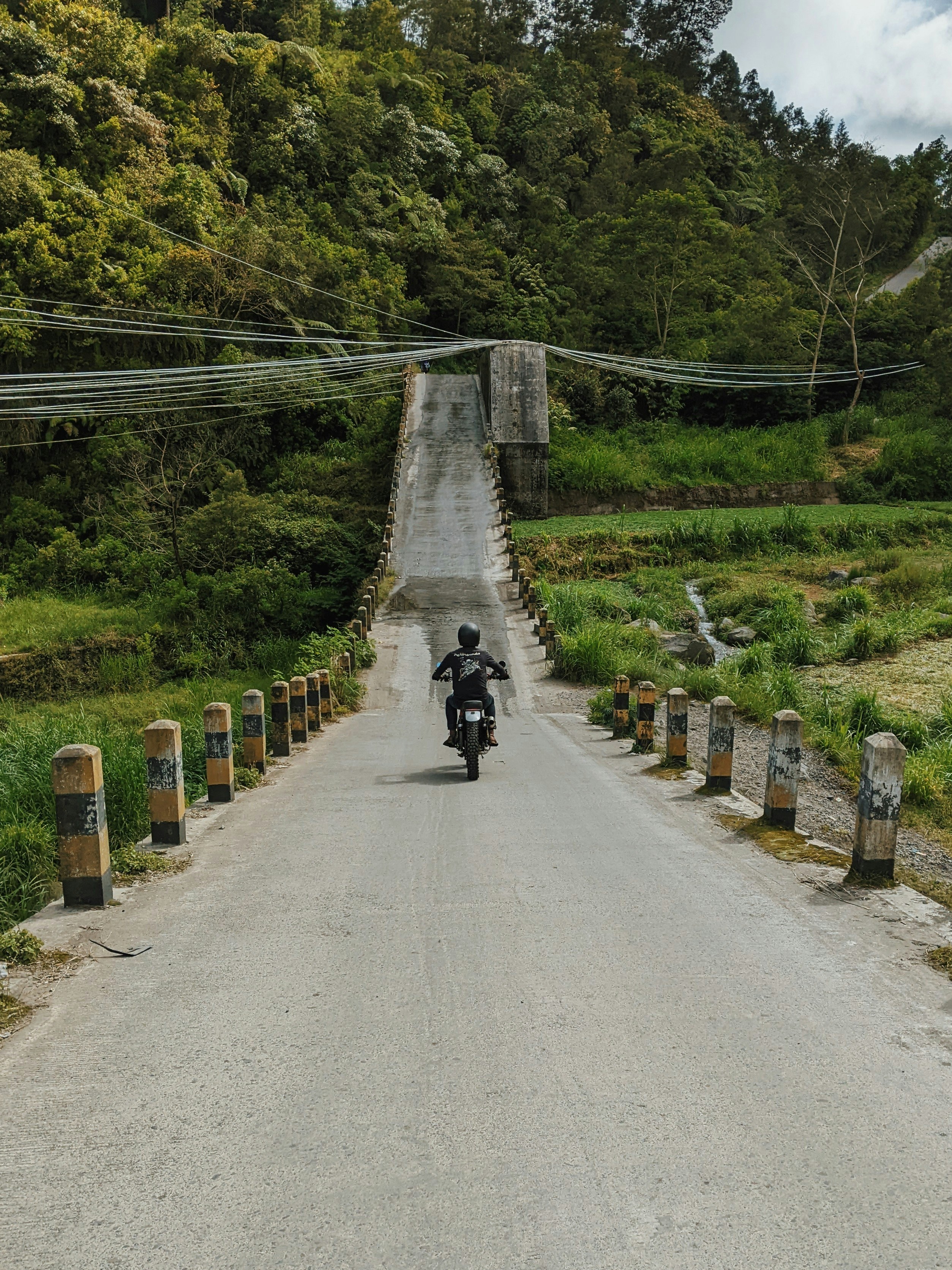 Motorcyclist crossing a narrow suspension bridge surrounded by lush greenery.