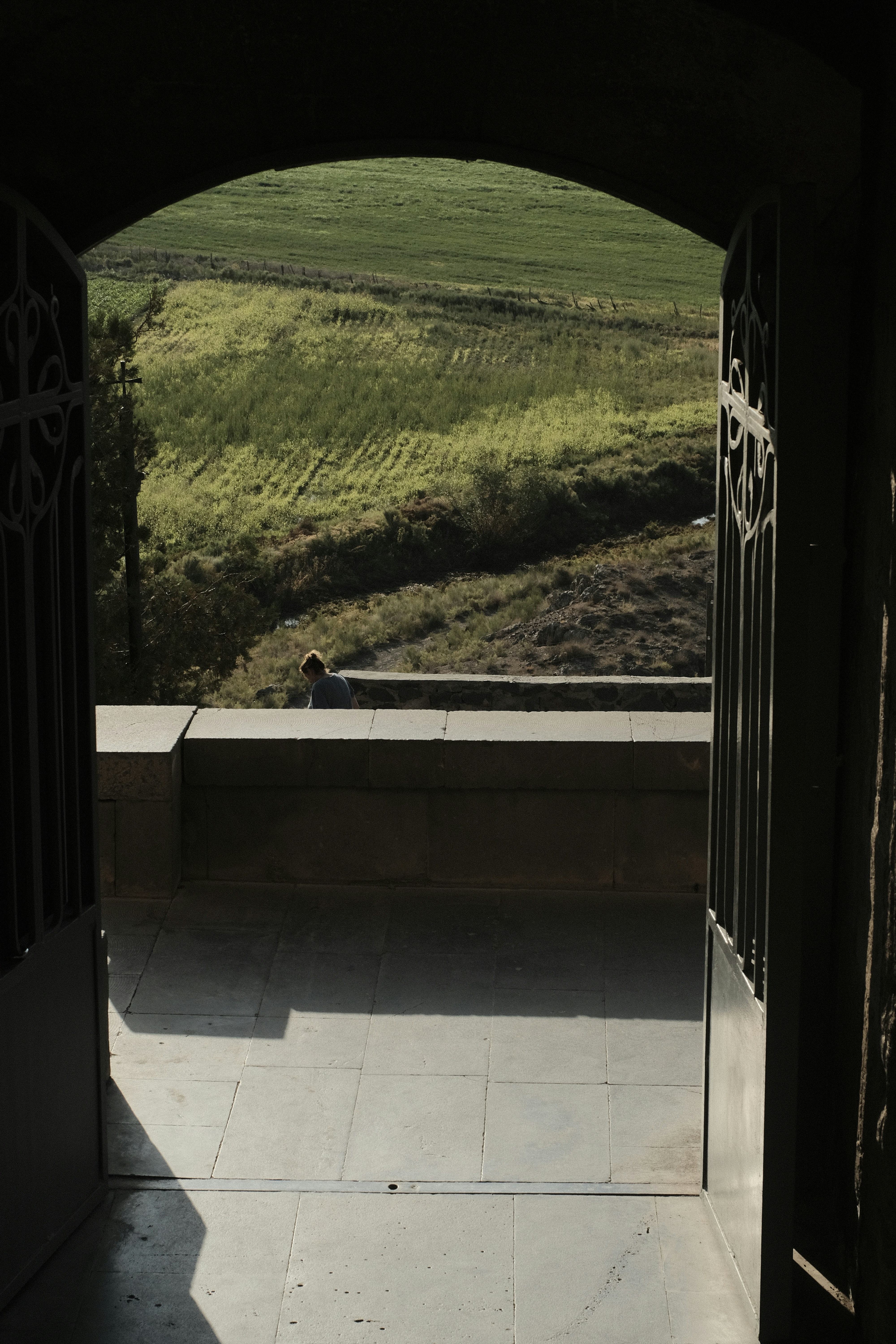 Threshold Light: A lone figure on a sunlit terrace beyond iron gates