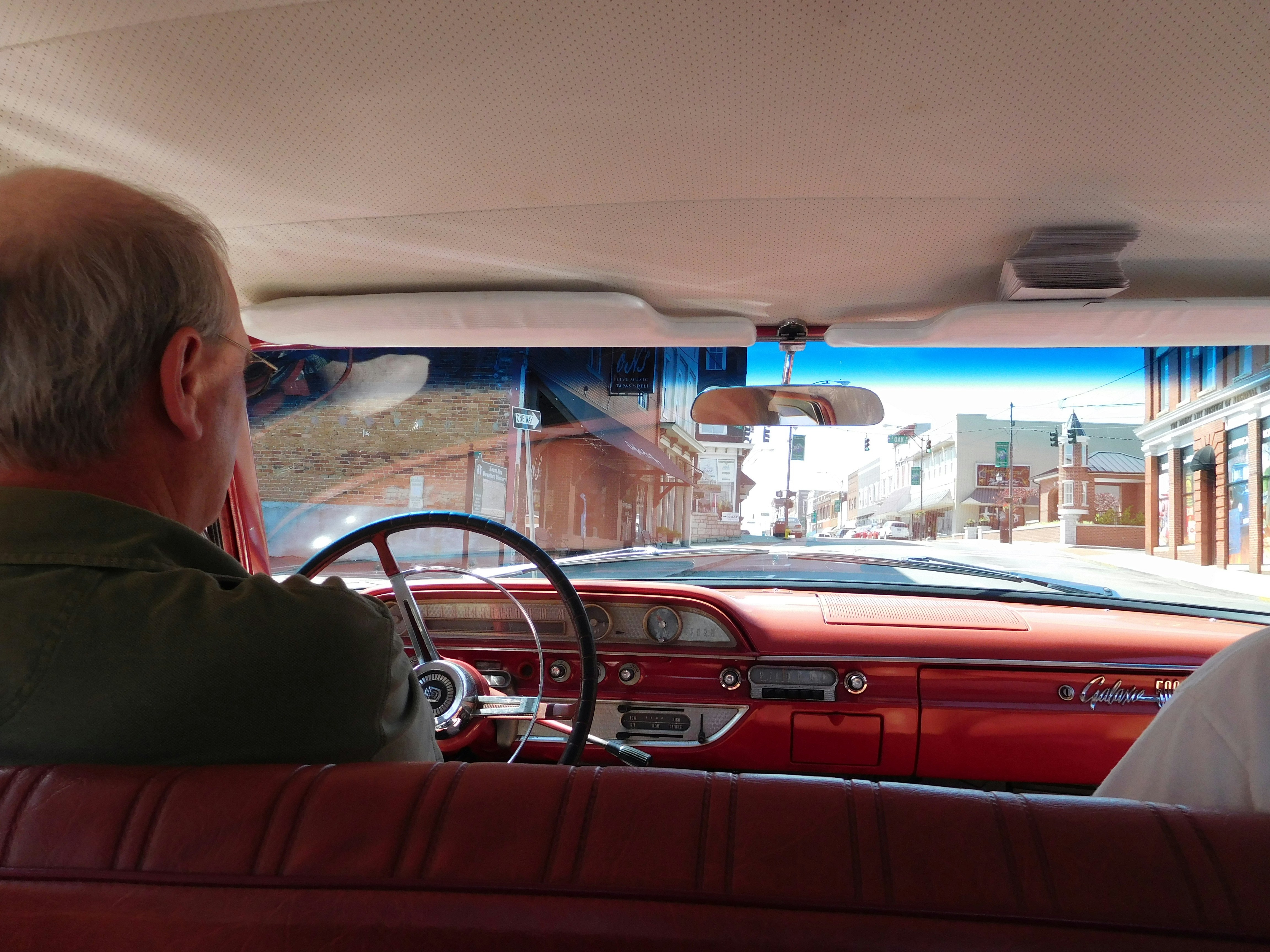 Interior view of a vintage red car with a chrome dashboard and steering wheel, a driver in the left seat, and a sunlit town street visible through the windshield.