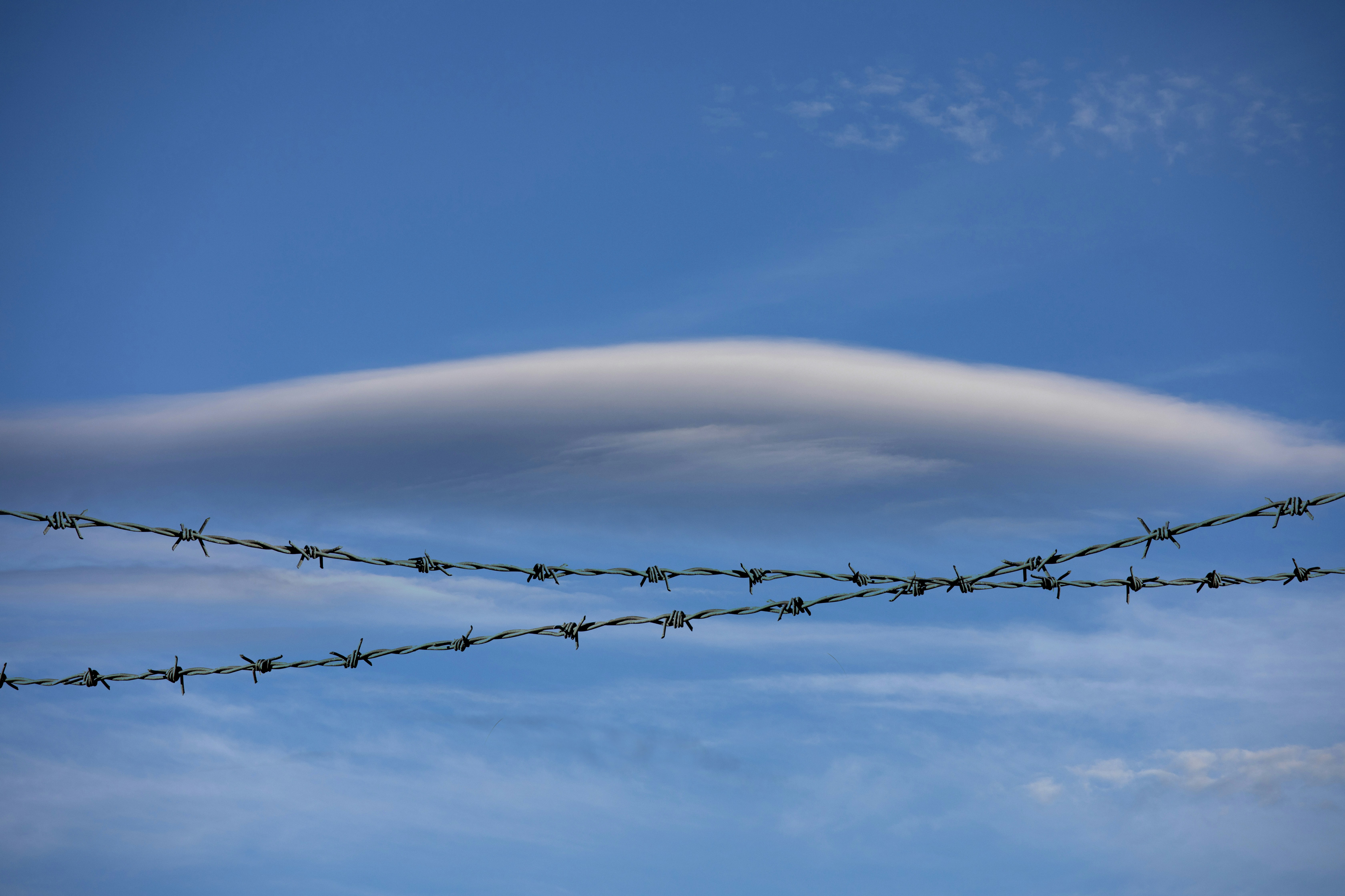 Barbed wire fence beneath a smooth, elongated cloud formation against a blue sky. The contrasting elements create a thought-provoking scene.