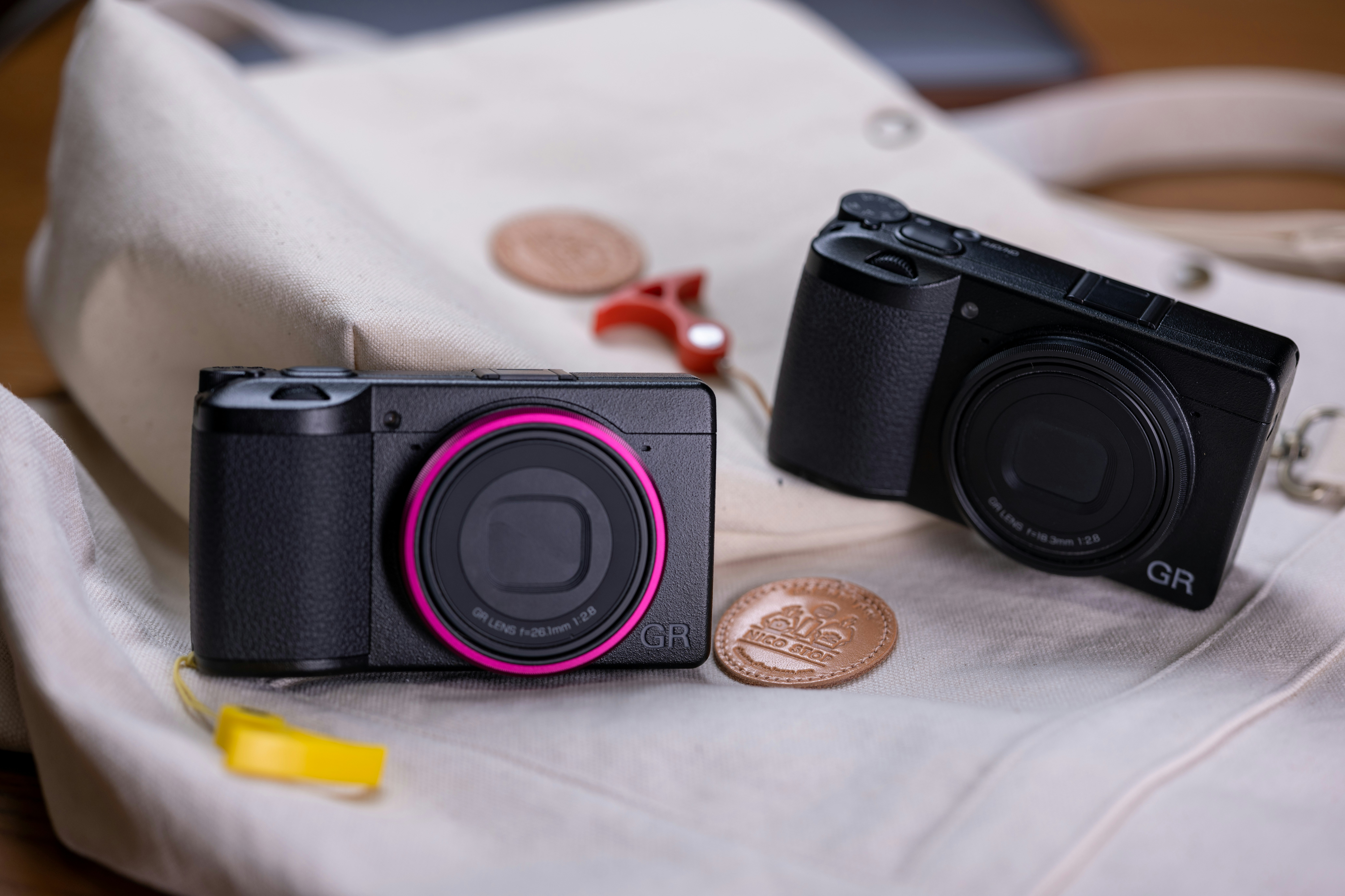Two compact cameras resting on a light fabric surface, accompanied by coins and a playful accessory. The scene showcases the blend of modern design and functionality.