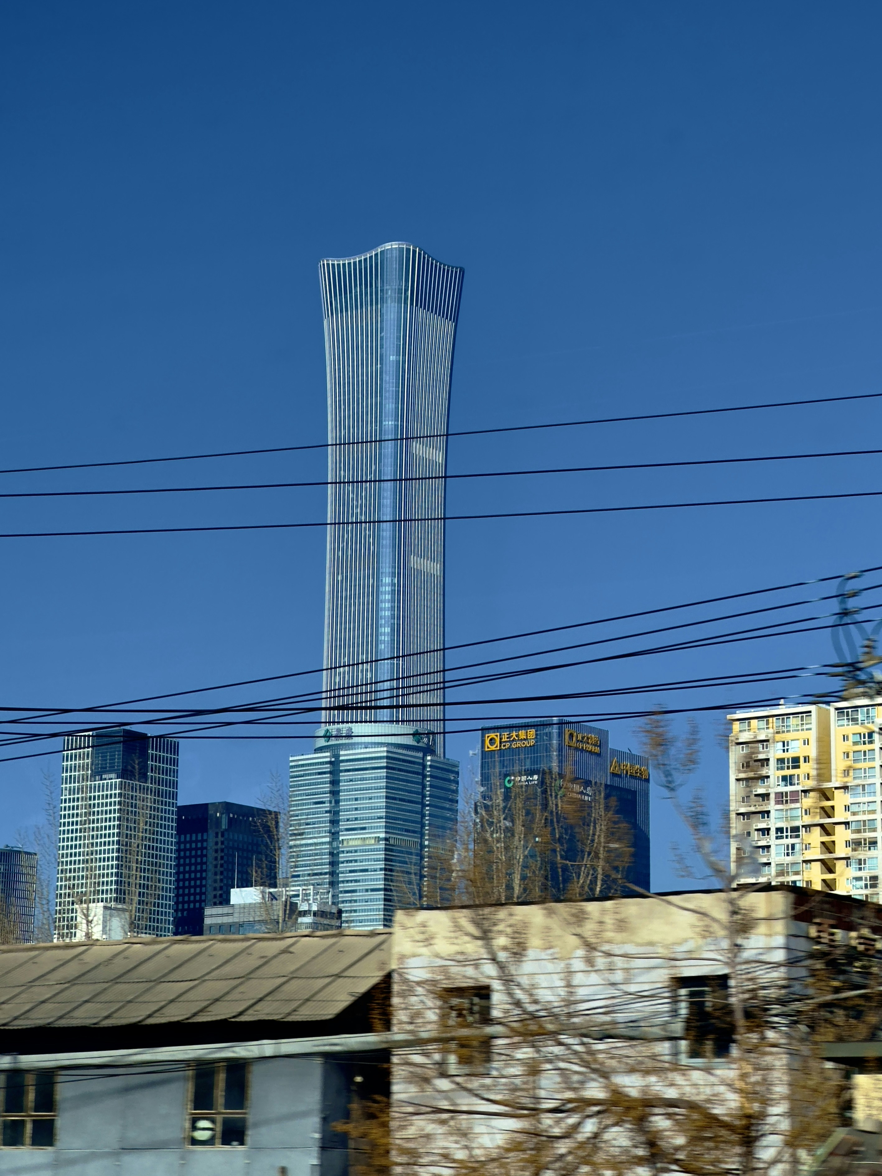 A sleek skyscraper rises against a clear blue sky, framed by urban buildings and power lines, showcasing contemporary architecture.