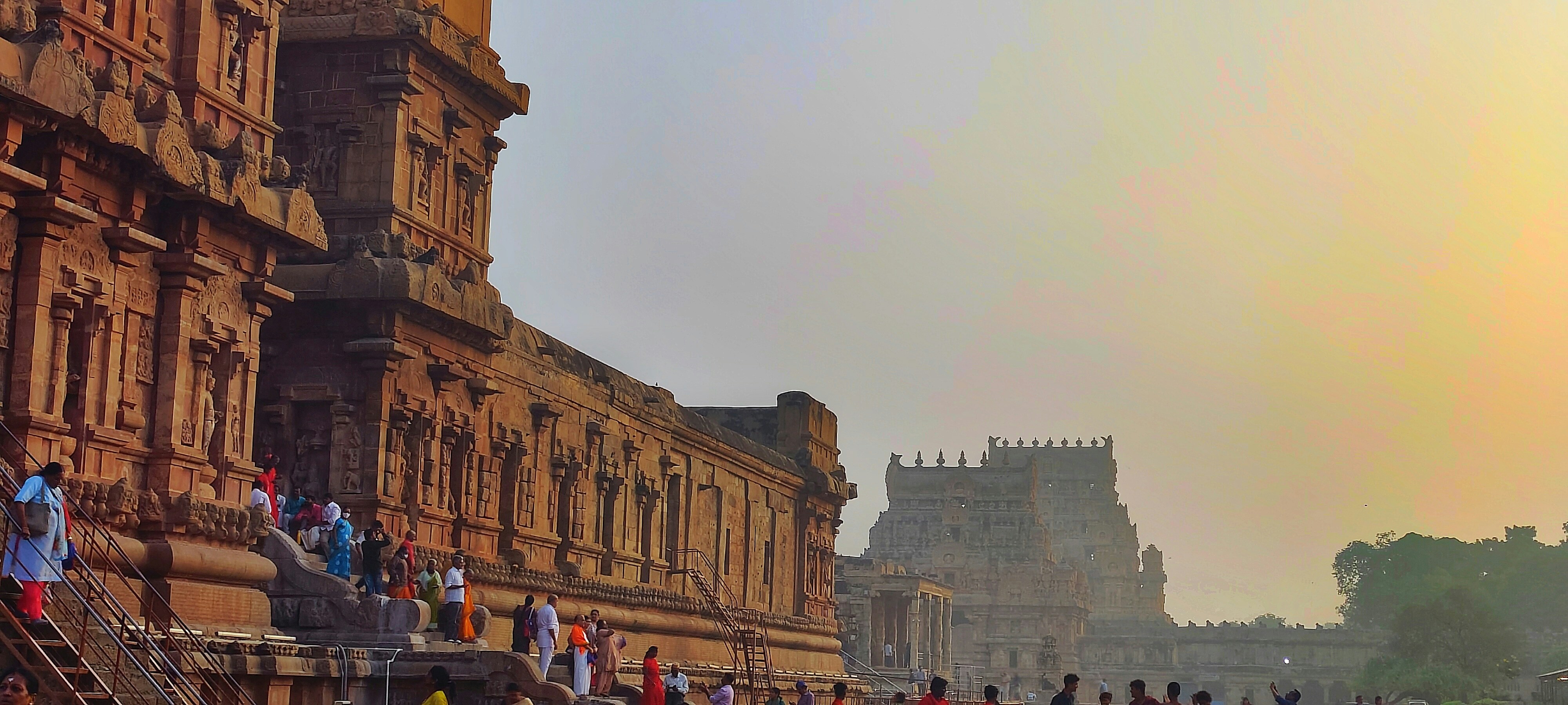 Majestic stone temples adorned with intricate carvings, bustling with visitors during the golden hour. The silhouette of a distant structure adds depth to the scene.
