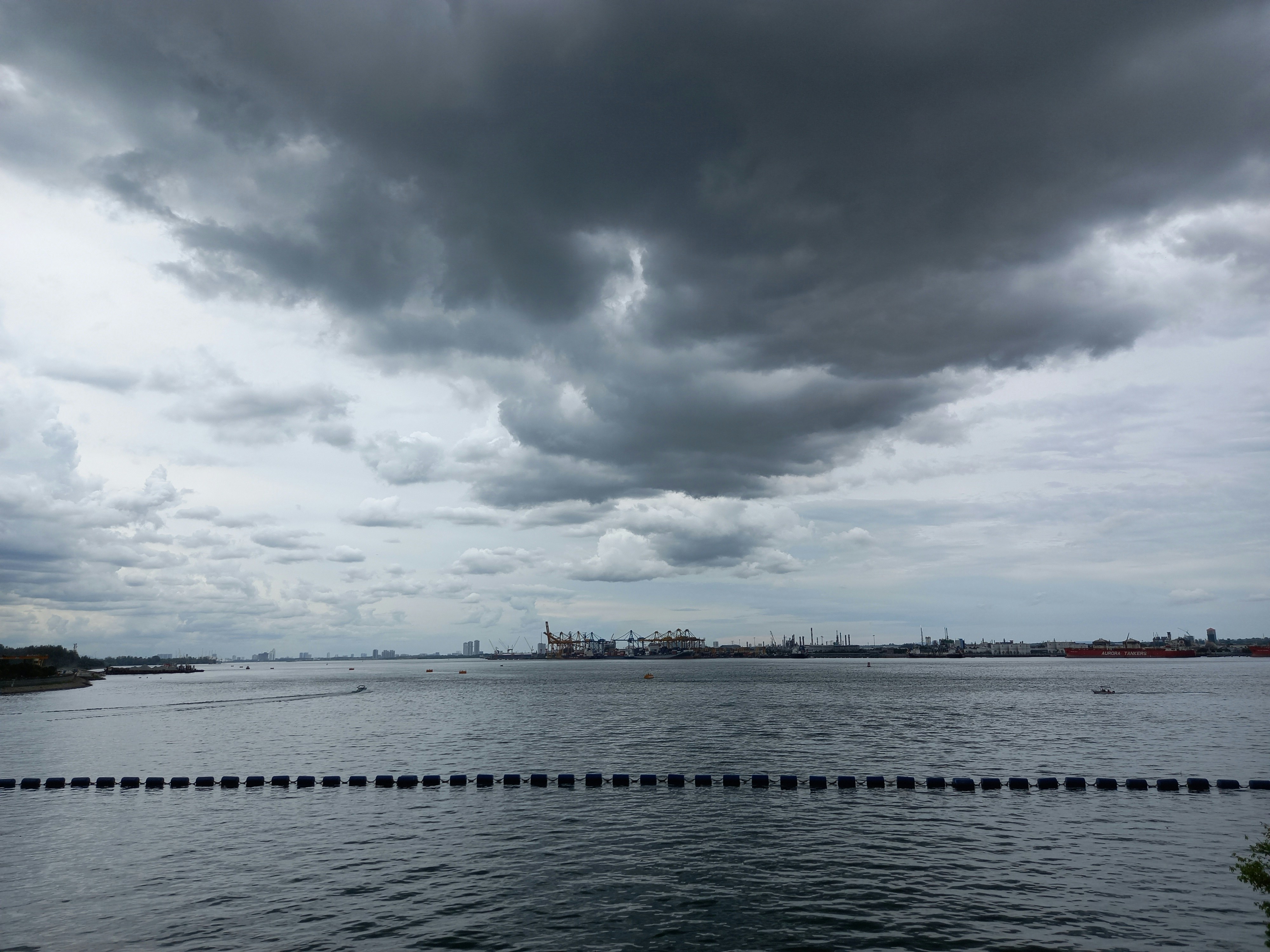 Storm clouds hover over a calm harbor, with a distant industrial skyline and a line of buoys crossing the water. This photograph emphasizes moody contrast and depth between sky and sea.