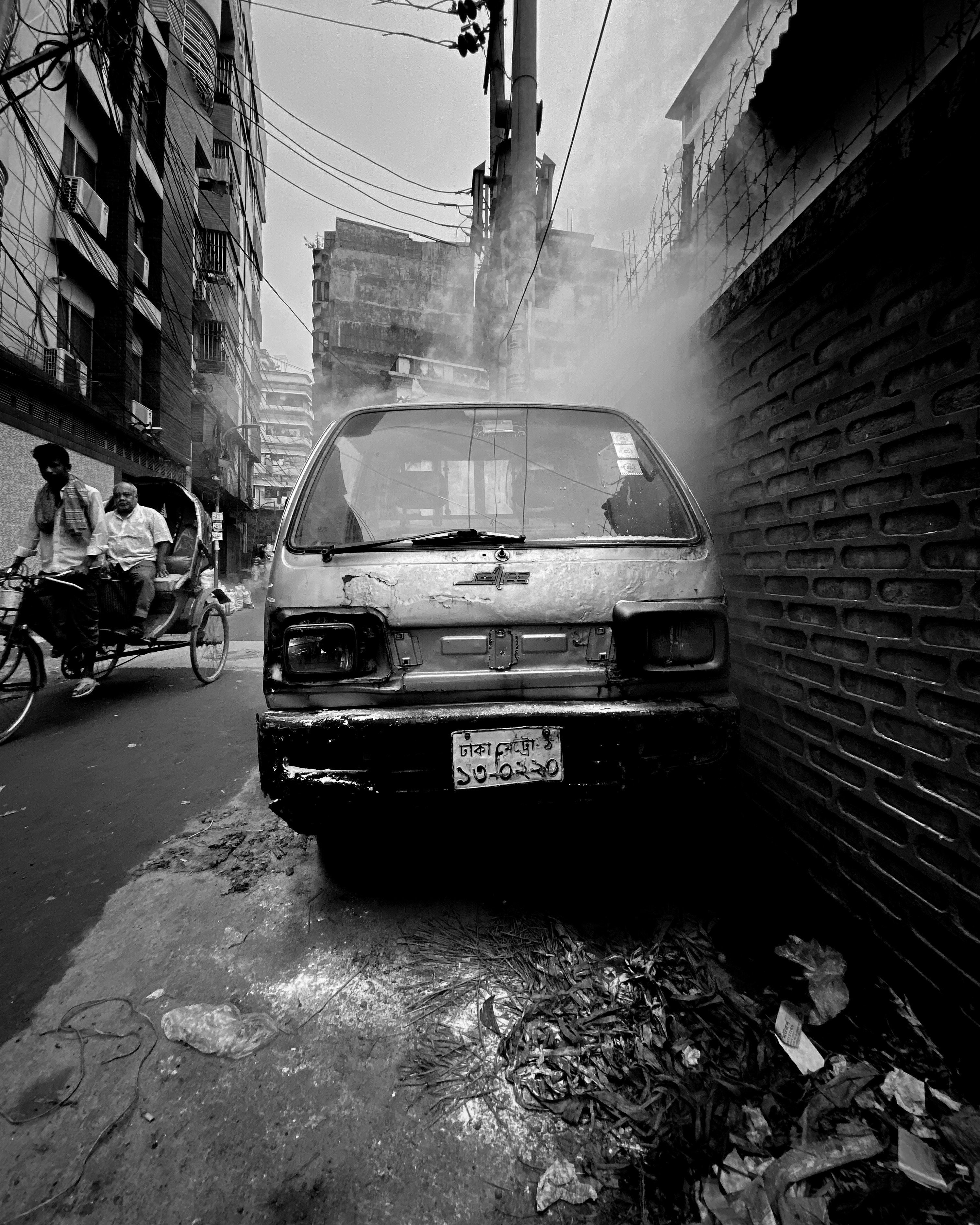 Abandoned car partially obscured by smoke, nestled against a brick wall in a busy urban street. Bicyclists pass by, highlighting the contrast between neglect and daily life.