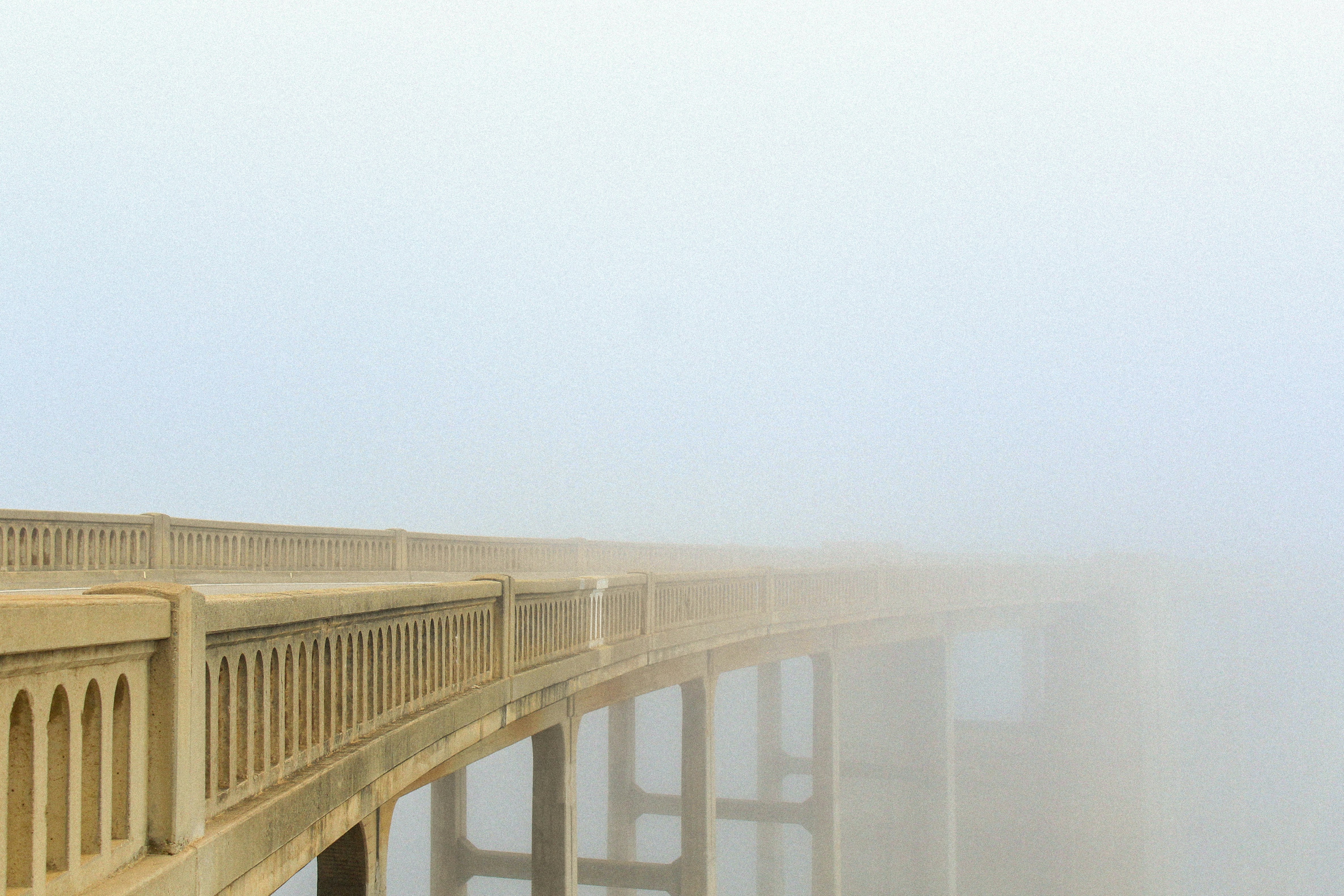 A winding bridge disappears into a thick fog, creating a sense of mystery and isolation. The architectural details are subtly obscured by the atmospheric conditions.
