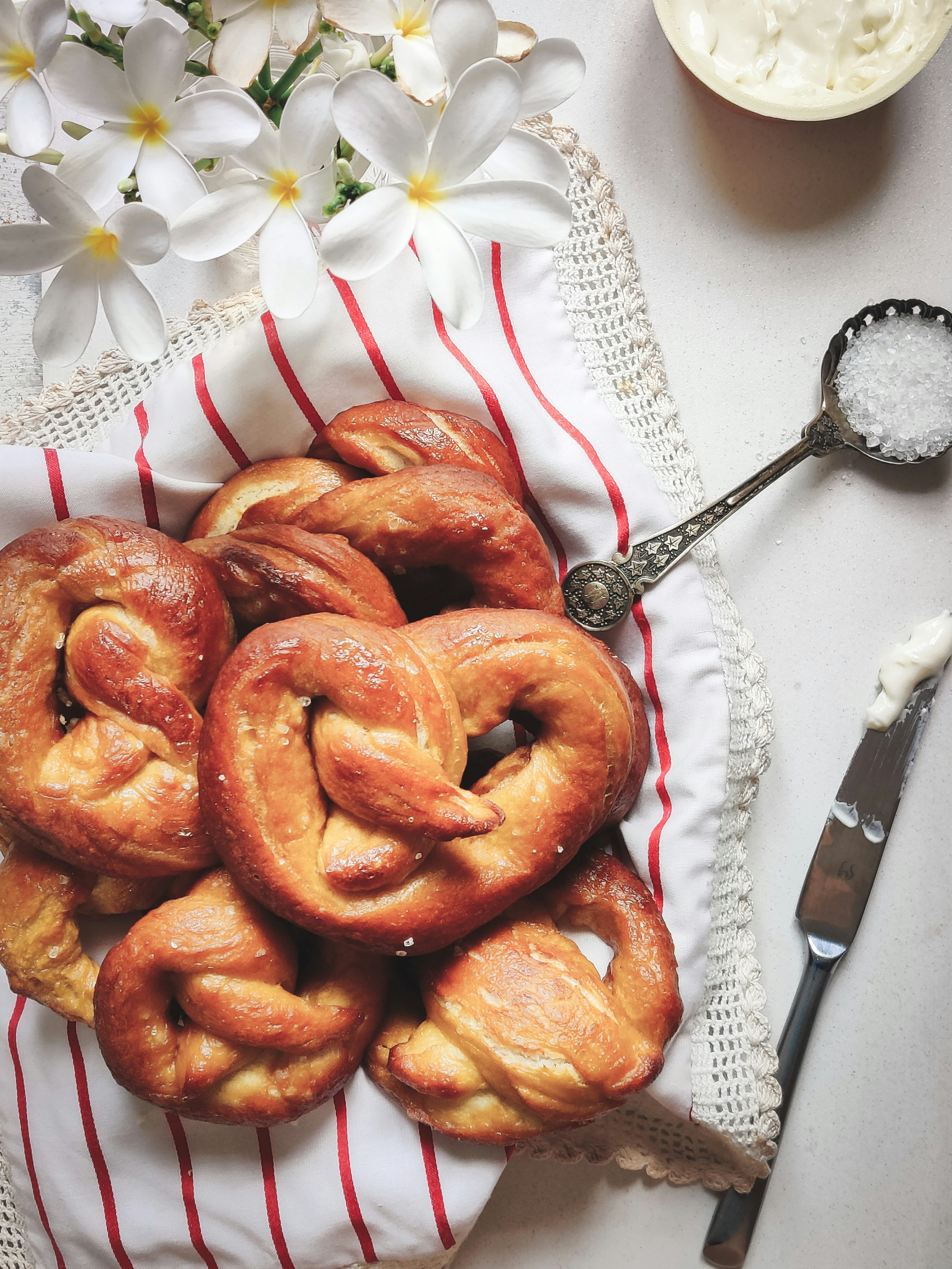 Golden-brown pretzels arranged on a striped cloth, accompanied by delicate flowers and a sprinkle of salt. A bowl of creamy spread sits nearby.
