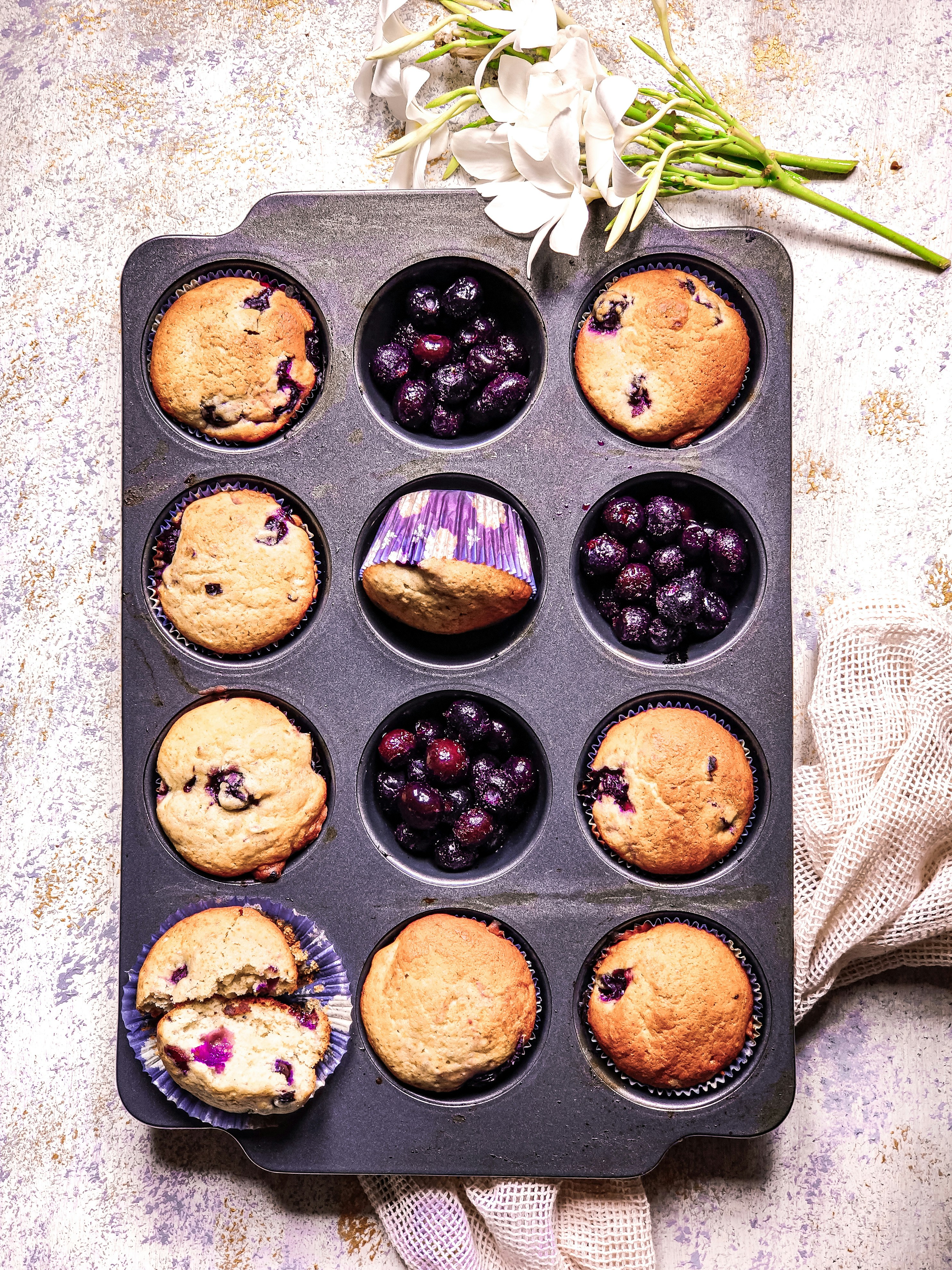 A photograph shows nine muffins in a muffin tin; the center muffin features purple frosting, surrounded by berry-loaded cups, with white flowers providing a delicate backdrop.