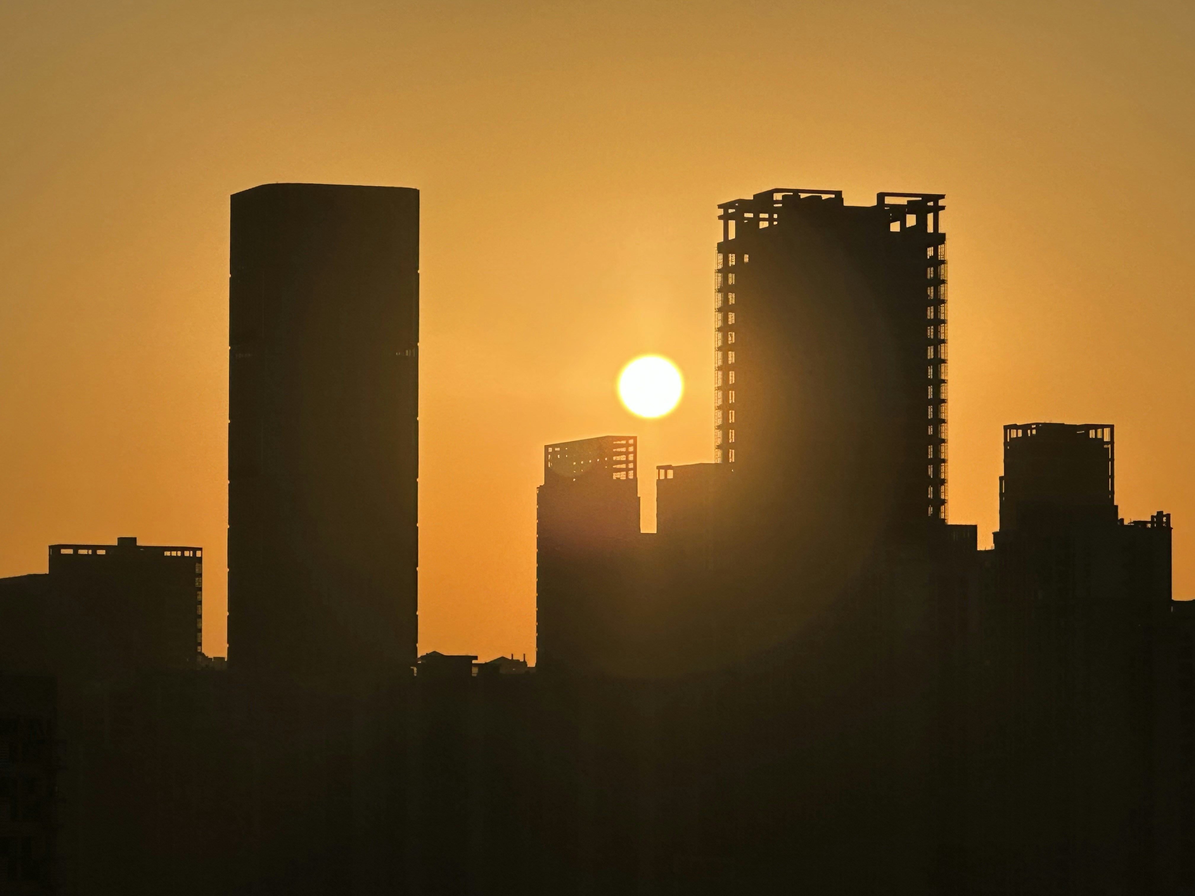 Silhouettes of skyscrapers against a vibrant sunset, with the sun positioned centrally behind the buildings.