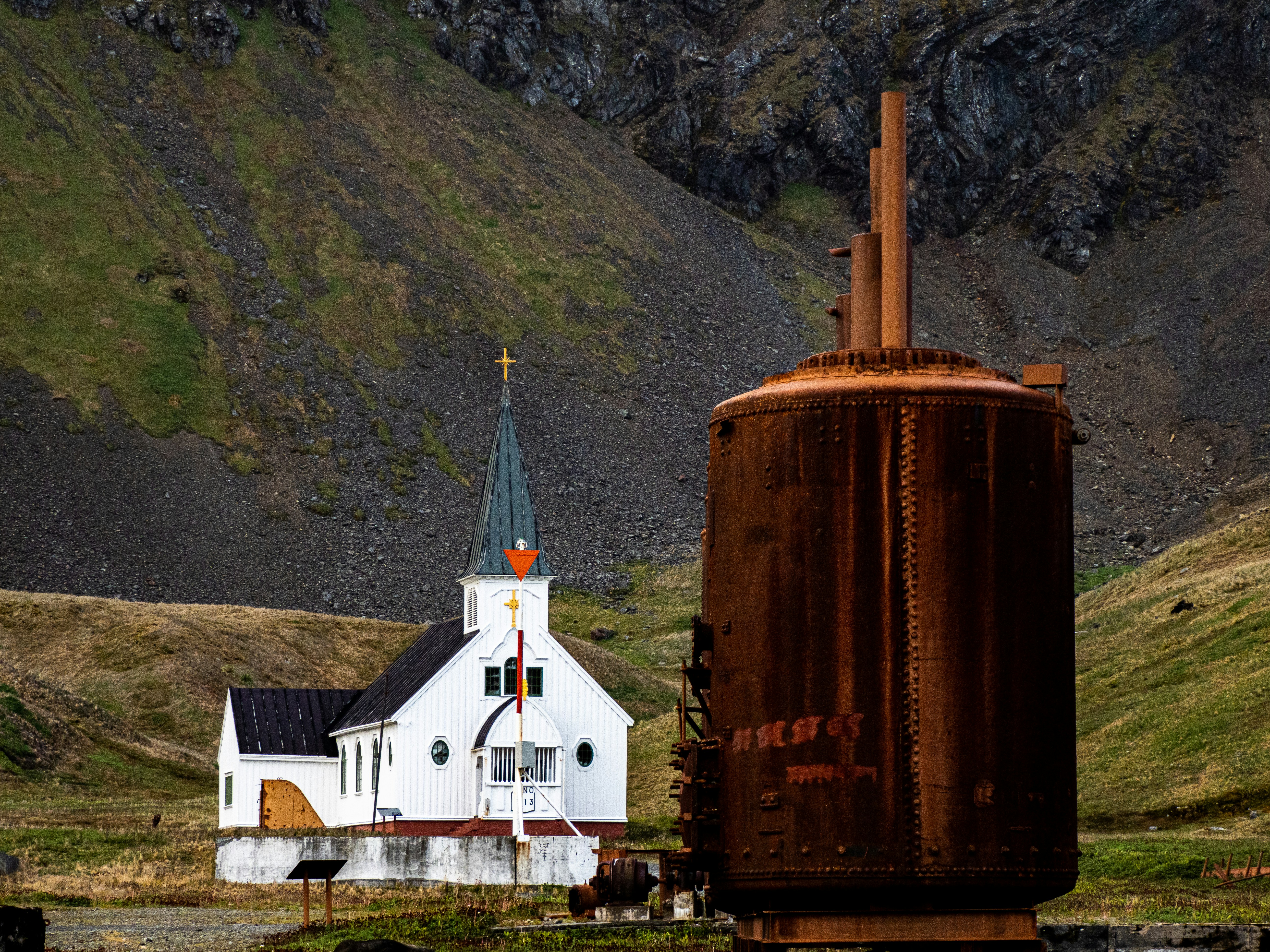 A quaint white church stands against rugged mountains, juxtaposed with a rusty industrial tank, illustrating the contrast between nature and human endeavor.