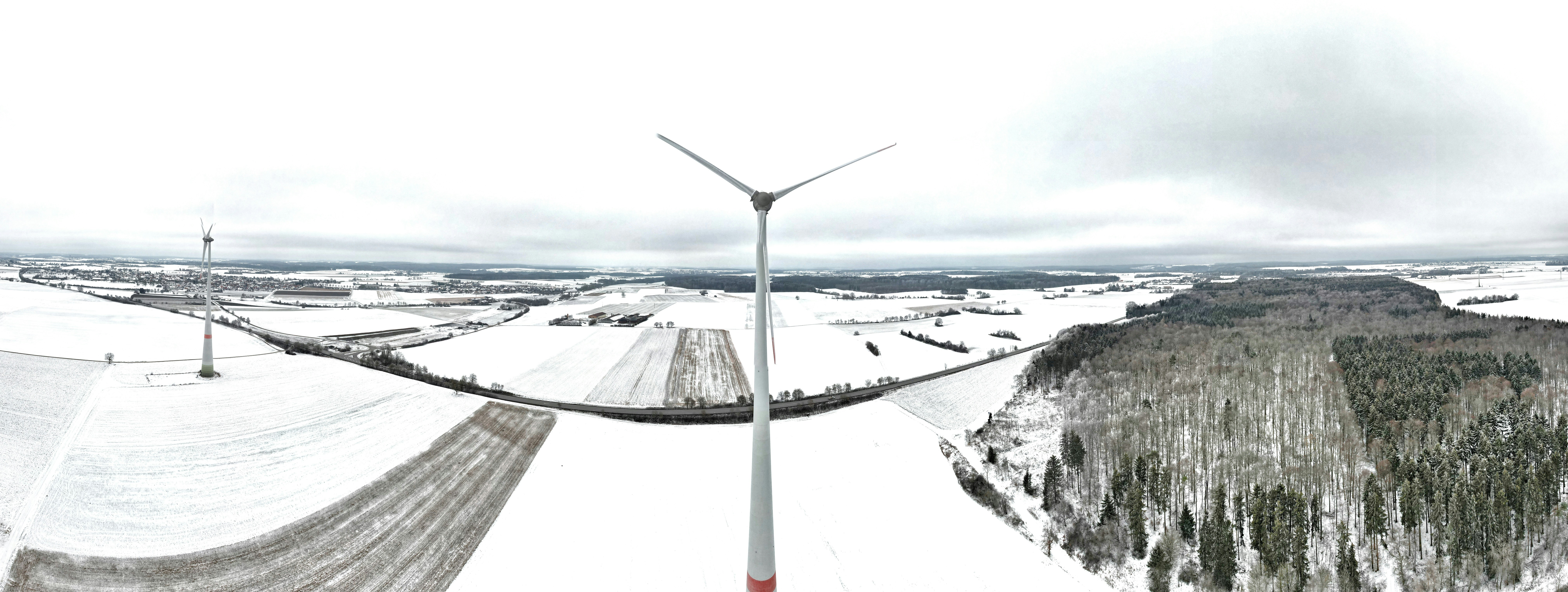 Panoramic aerial view of snow-blanketed fields and a wind turbine under cloudy skies.