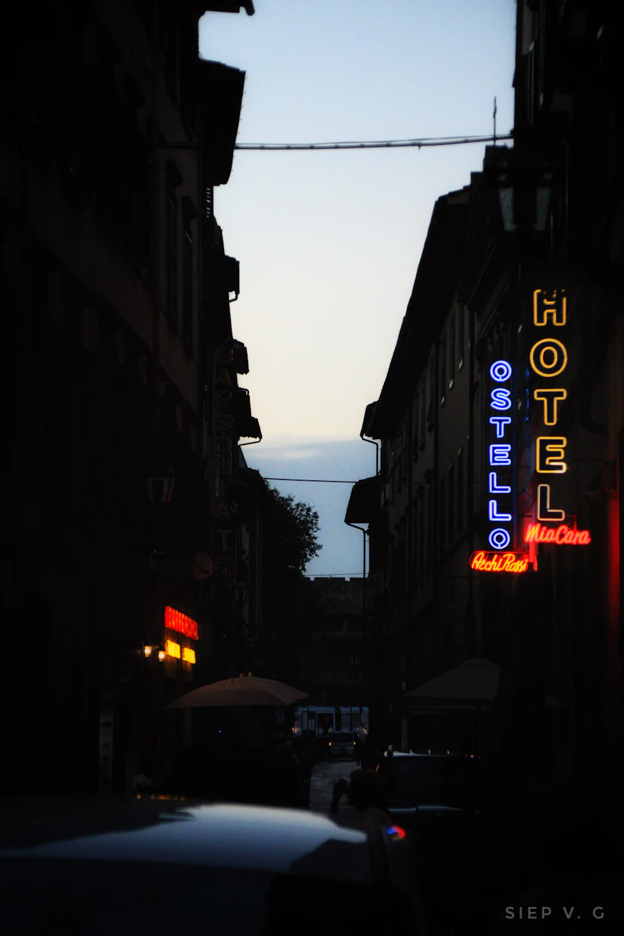 Dark narrow street framed by tall buildings at dusk, with glowing vertical hotel neon signs on the right and a pale sky overhead. A few umbrellas and parked cars sit in the shadowed foreground.