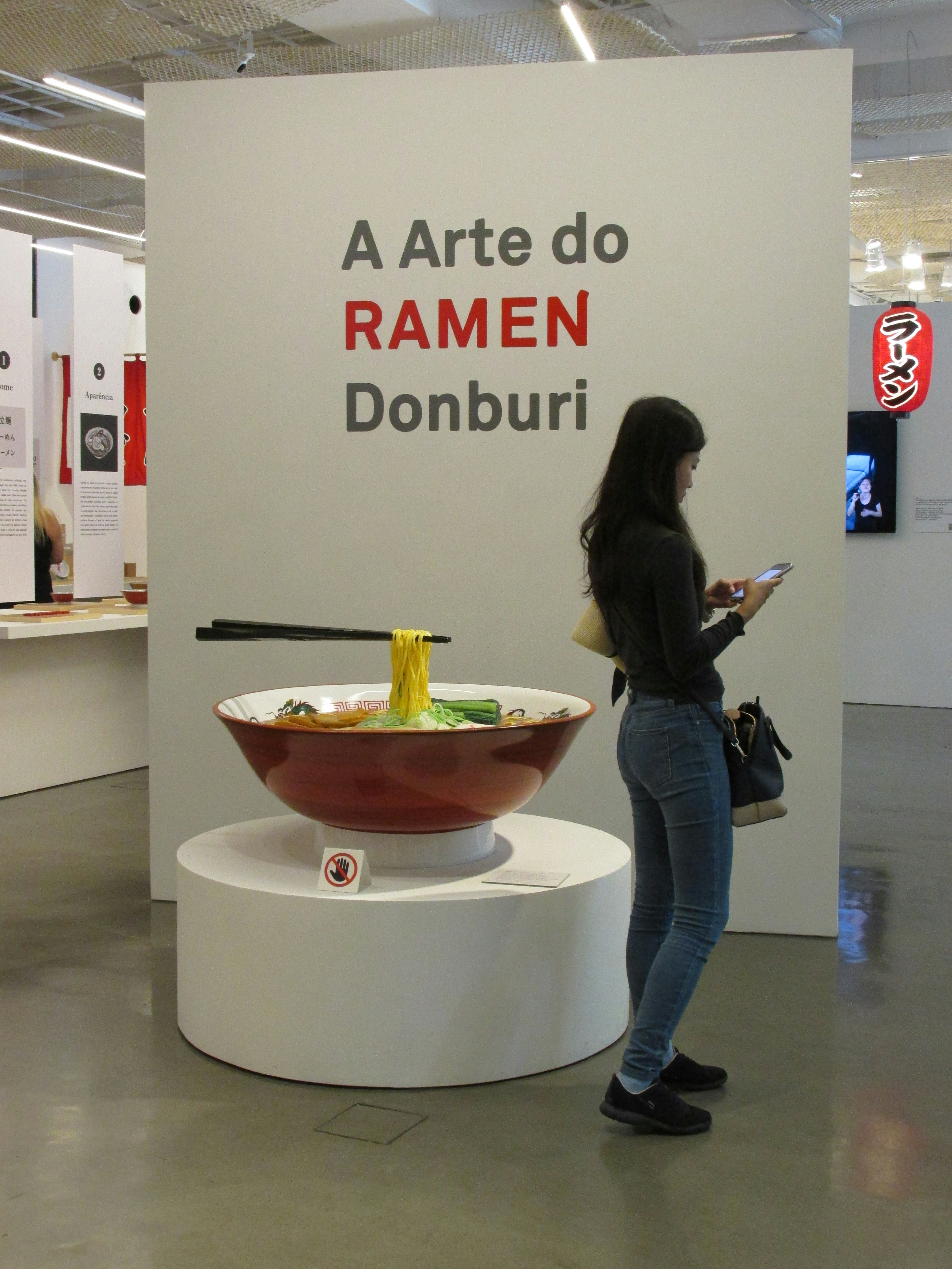 A large ramen bowl sculpture on a white pedestal dominates a contemporary gallery, with a woman nearby looking at her phone.