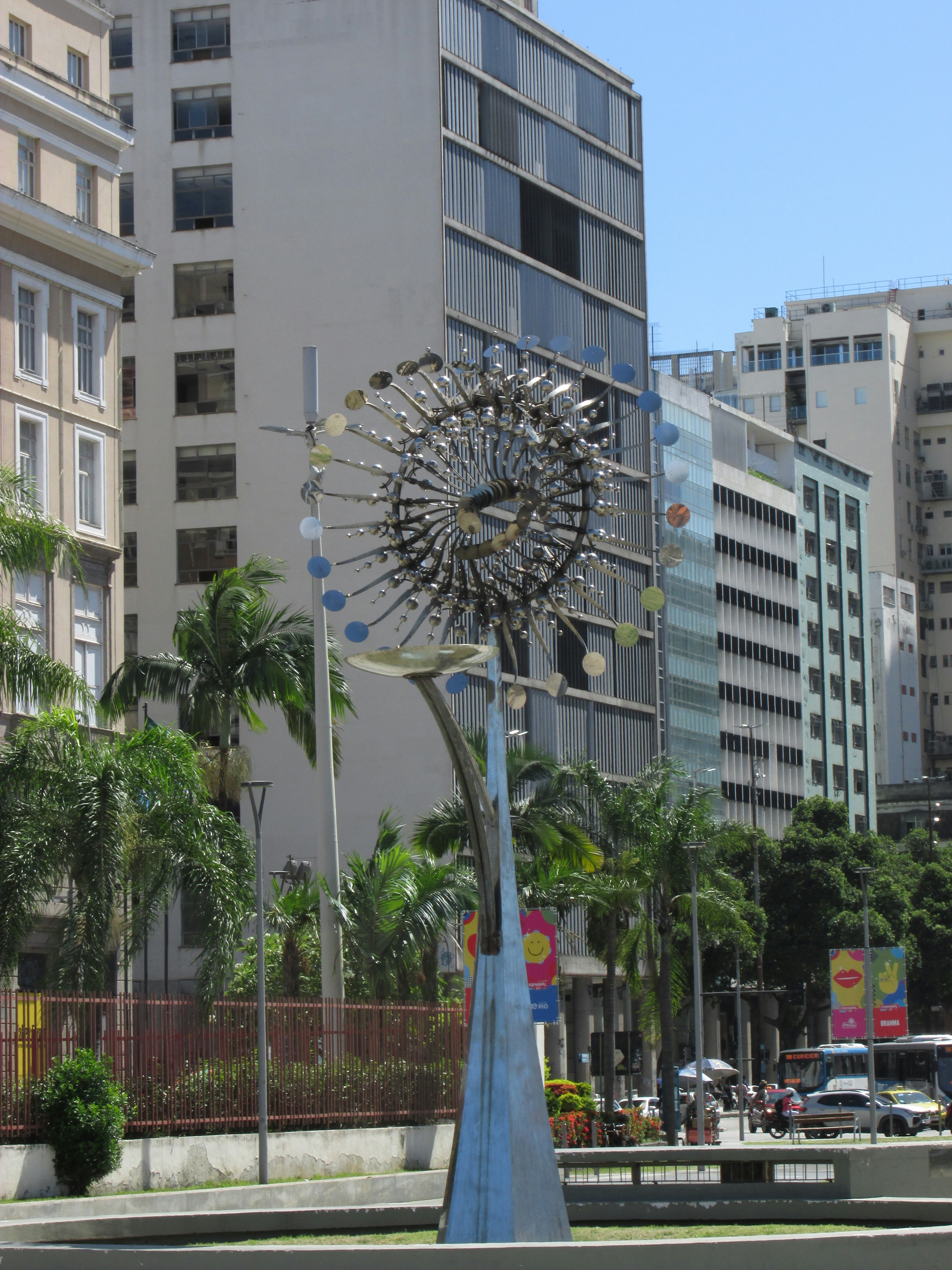 A contemporary sculpture featuring a circular design with colorful elements, set against a backdrop of modern architecture and palm trees.
