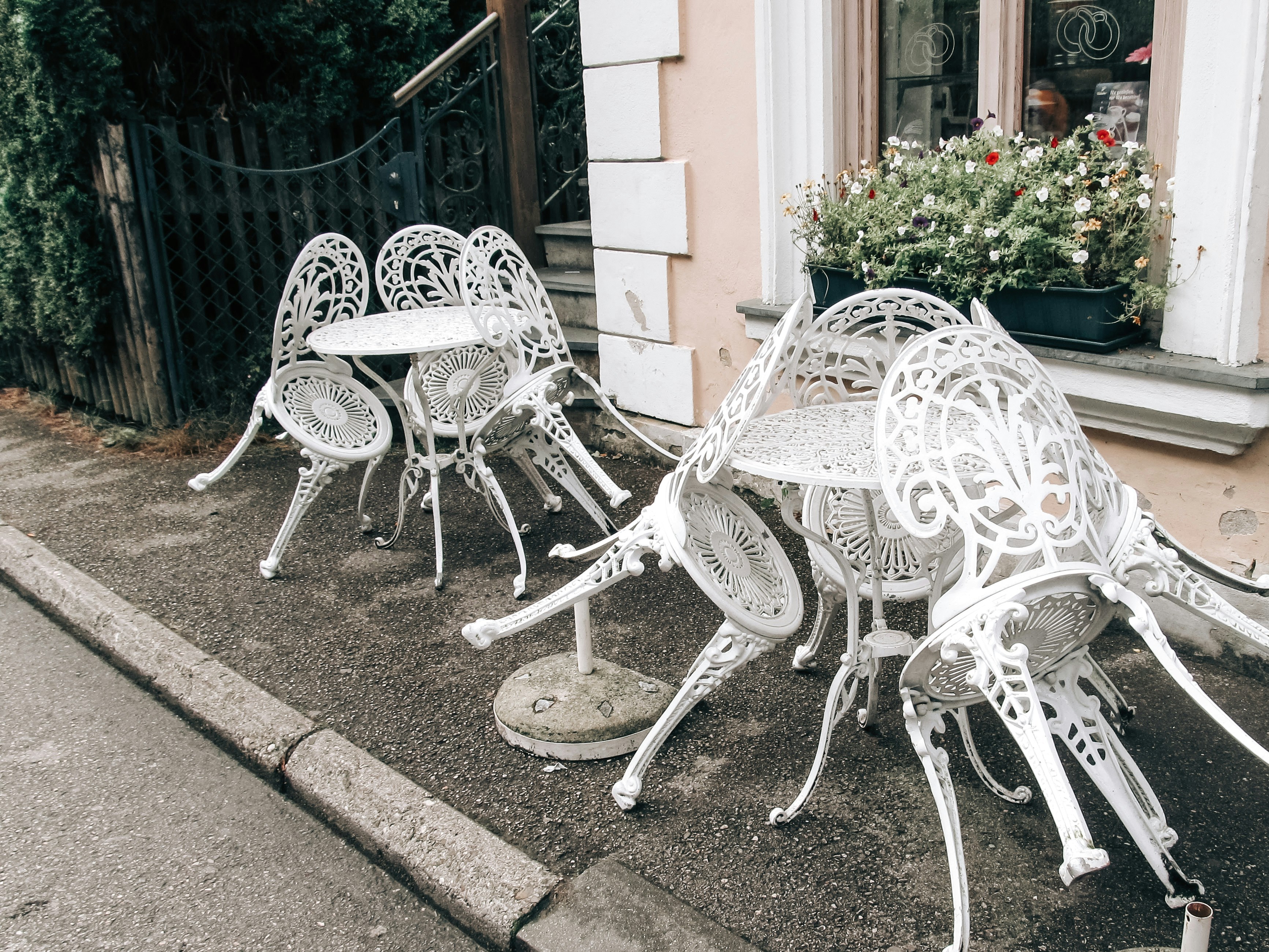 White wrought-iron cafe tables and chairs spill along a curb on a quiet street, their intricate patterns catching the light. A storefront with flower-filled window boxes provides a soft backdrop.