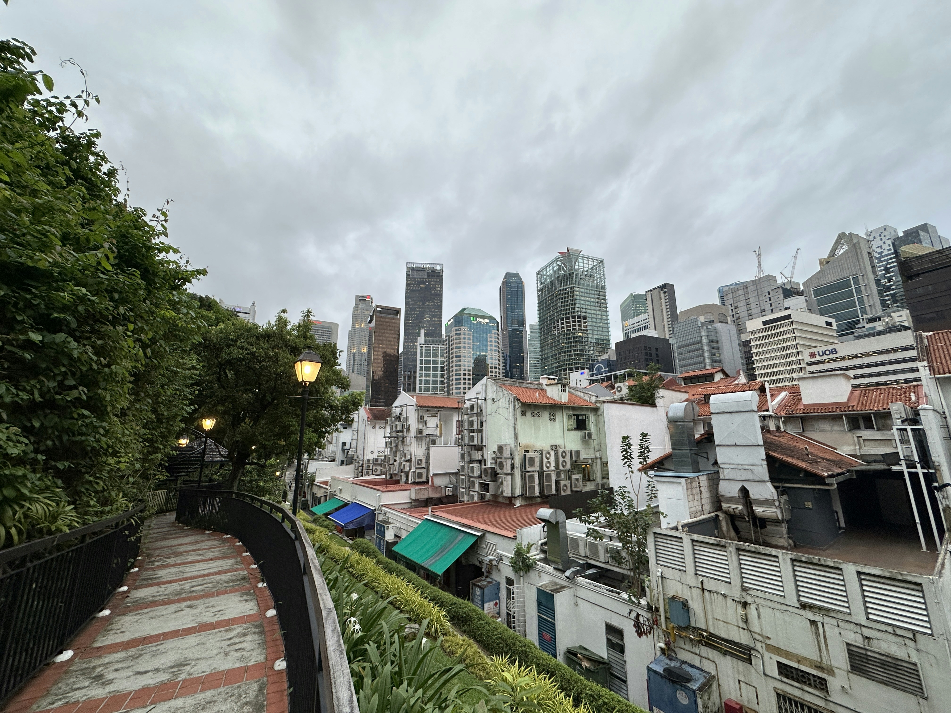 A lush pathway lined with greenery leads to a vibrant cityscape, showcasing a blend of historical architecture and modern skyscrapers under a cloudy sky.