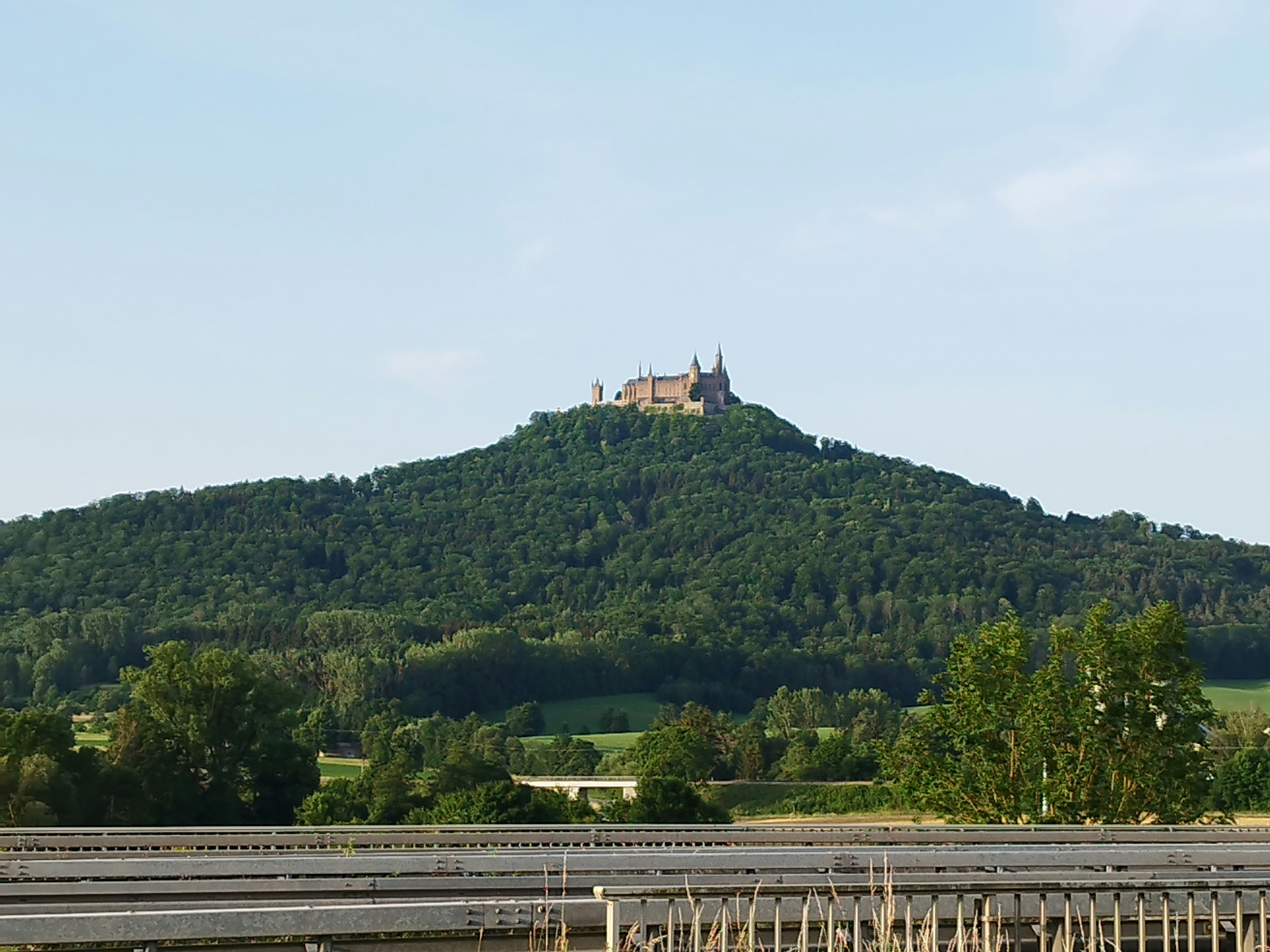 A medieval castle perched atop a lush green hill, surrounded by sprawling fields and trees under a clear sky.