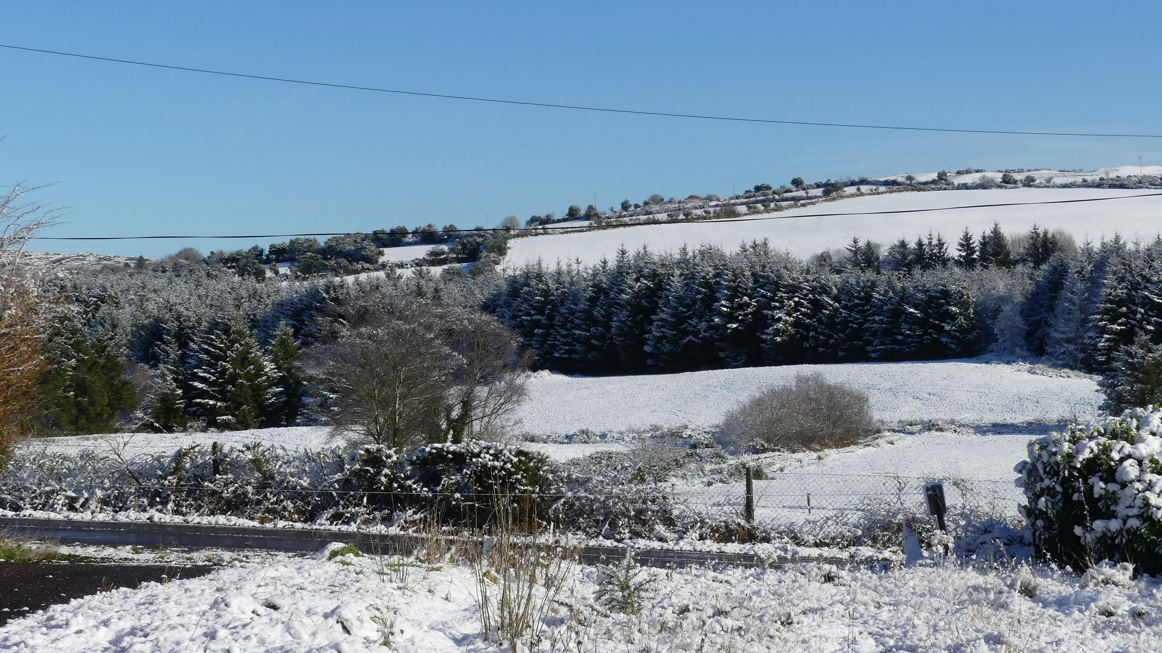 Snow-covered hills and evergreen trees under a clear blue sky create a serene winter scene. The landscape captures the tranquil beauty of nature in its frosty attire.