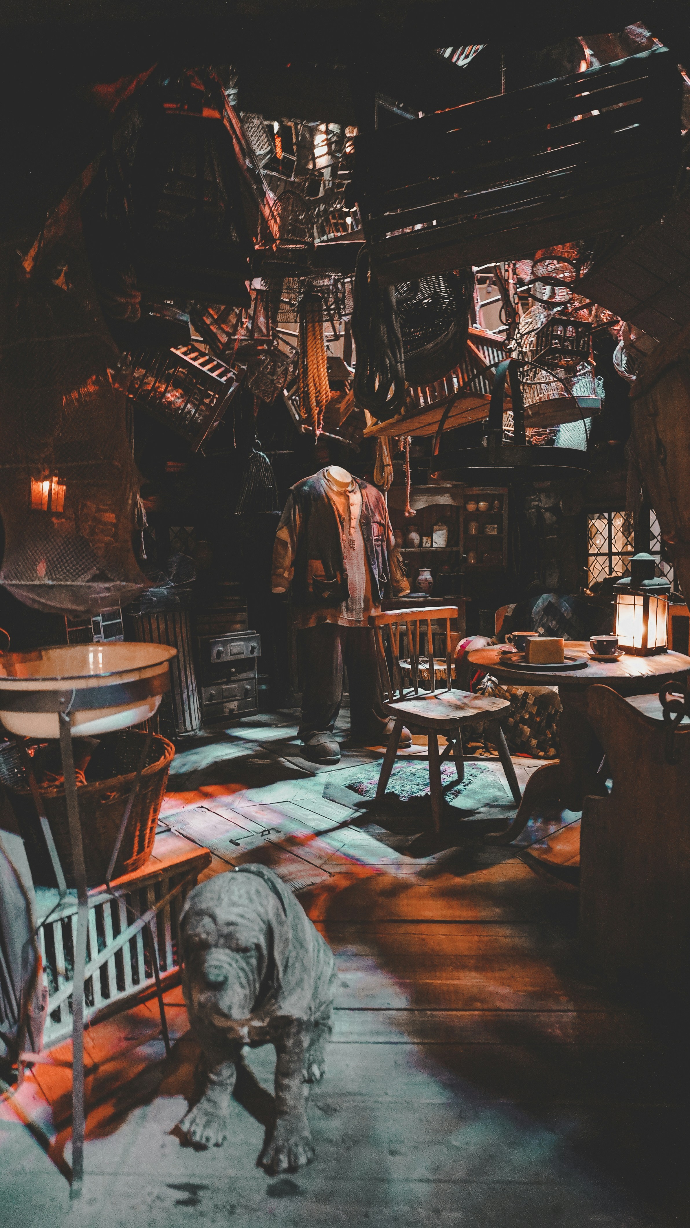 Dimly lit, cluttered interior with hanging objects and a stoic mannequin in the center, a dog at the foreground, and a warm table lamp casting amber light.