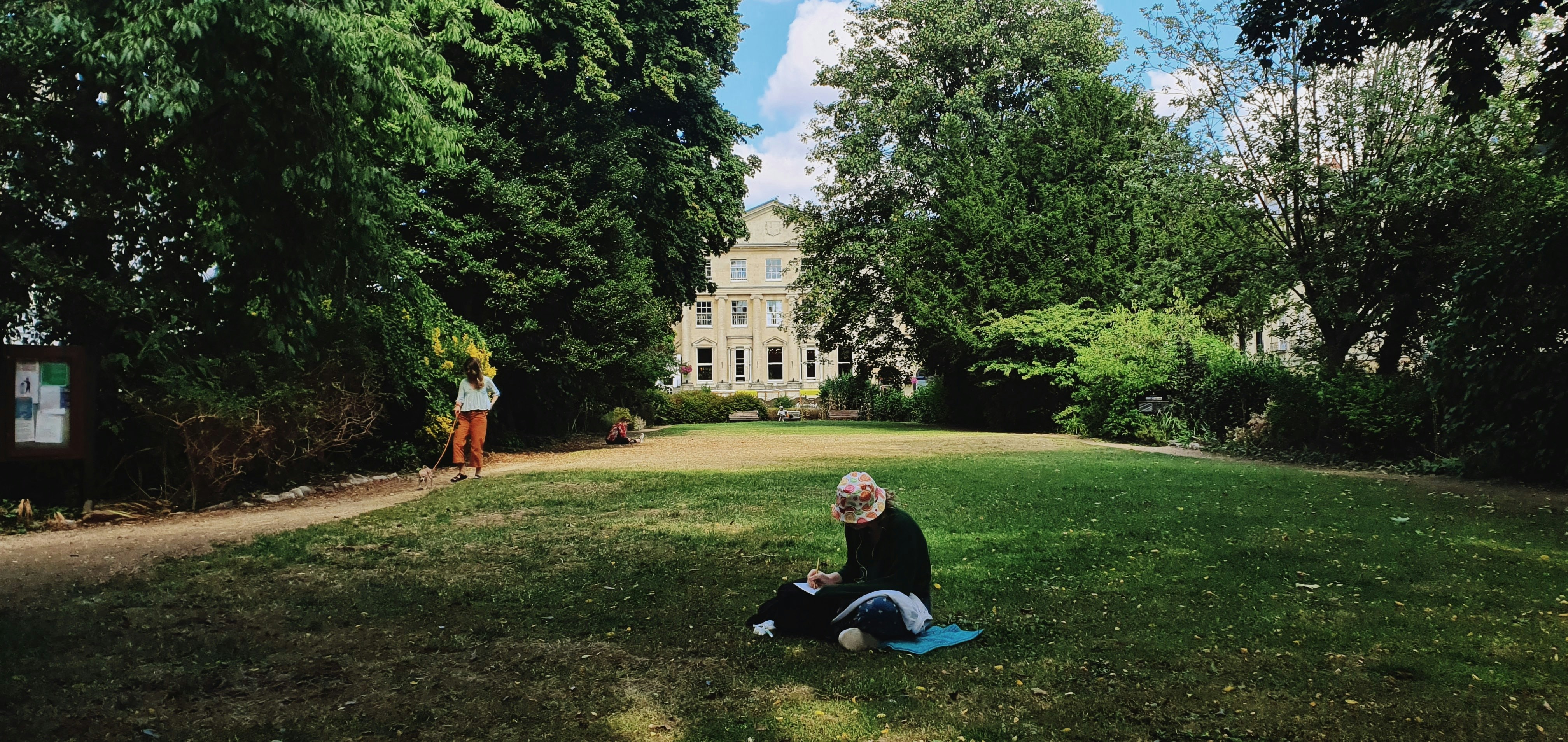 Person sitting on grass in a serene park surrounded by lush trees and distant buildings under a blue sky.