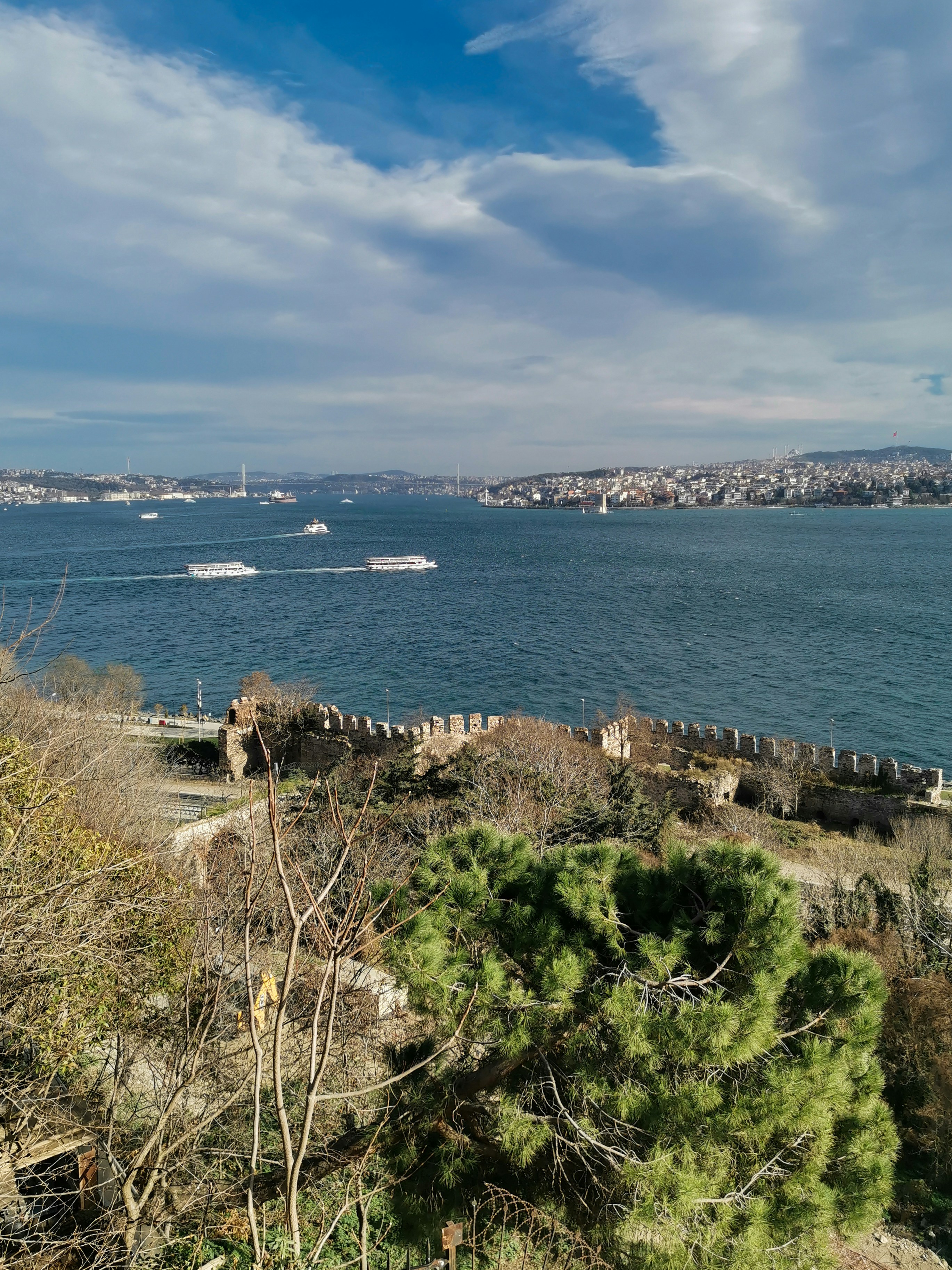 View of boats navigating the Bosphorus Strait, framed by lush greenery and ancient fortifications. The skyline of Istanbul rises in the background.