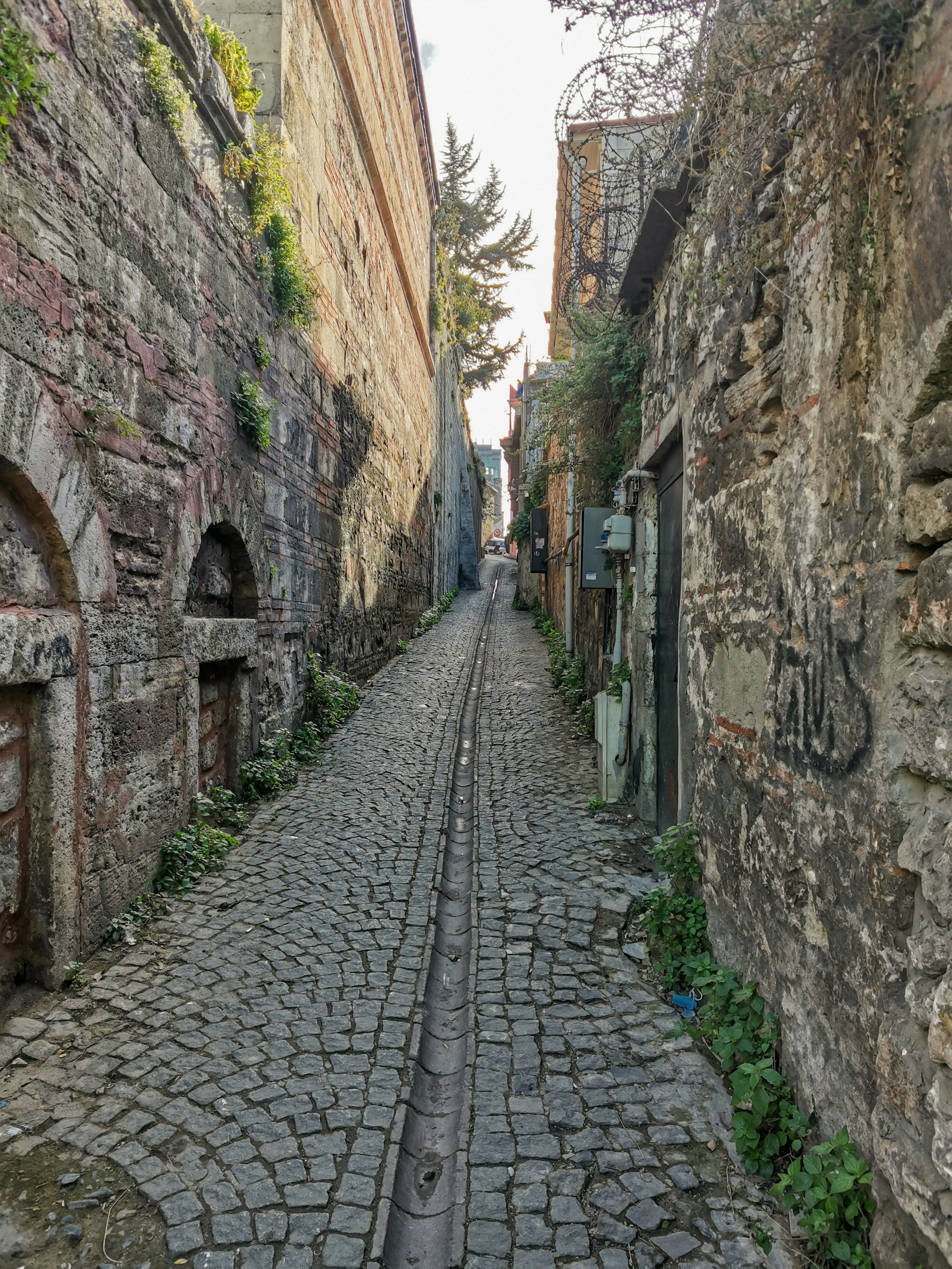 Photograph of a narrow cobblestone alley flanked by weathered stone walls, stretching toward a distant light. The scene emphasizes textural detail and perspective along the central gutter.