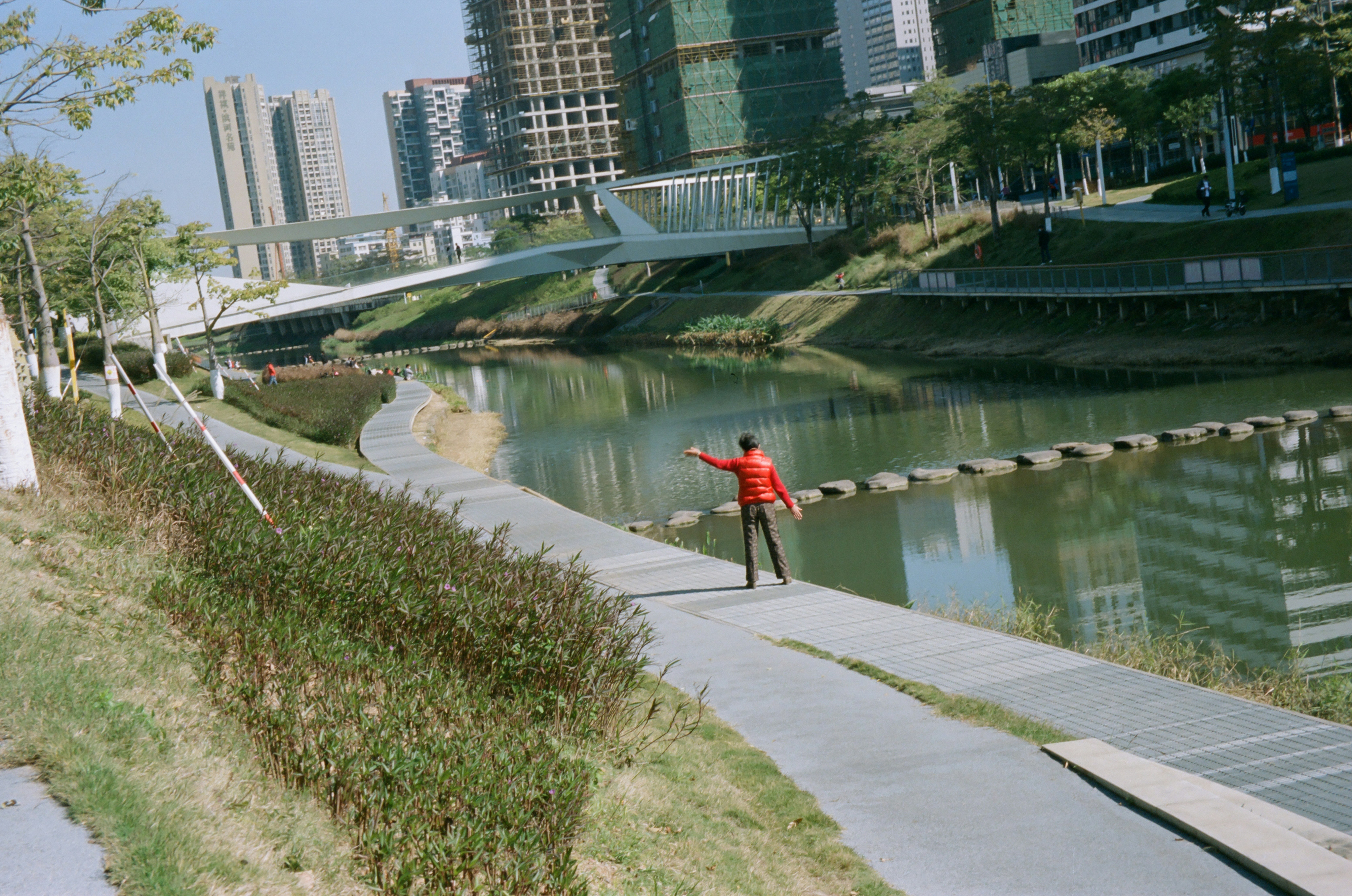 Red-clad figure anchors a sunlit riverside promenade beneath a curving city bridge